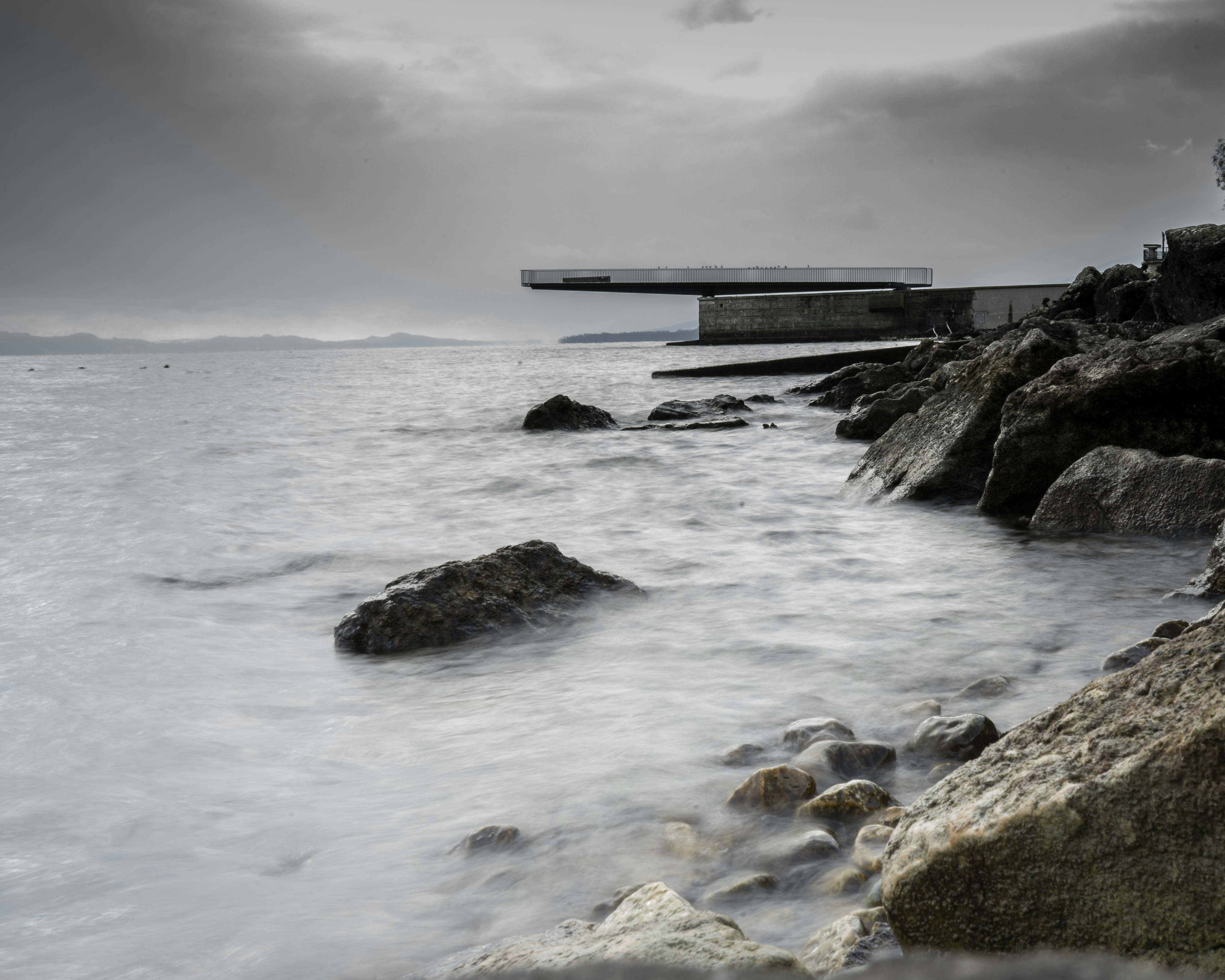 Grayscale scene of rocky coastline meeting a calm sea under a cloudy sky.
