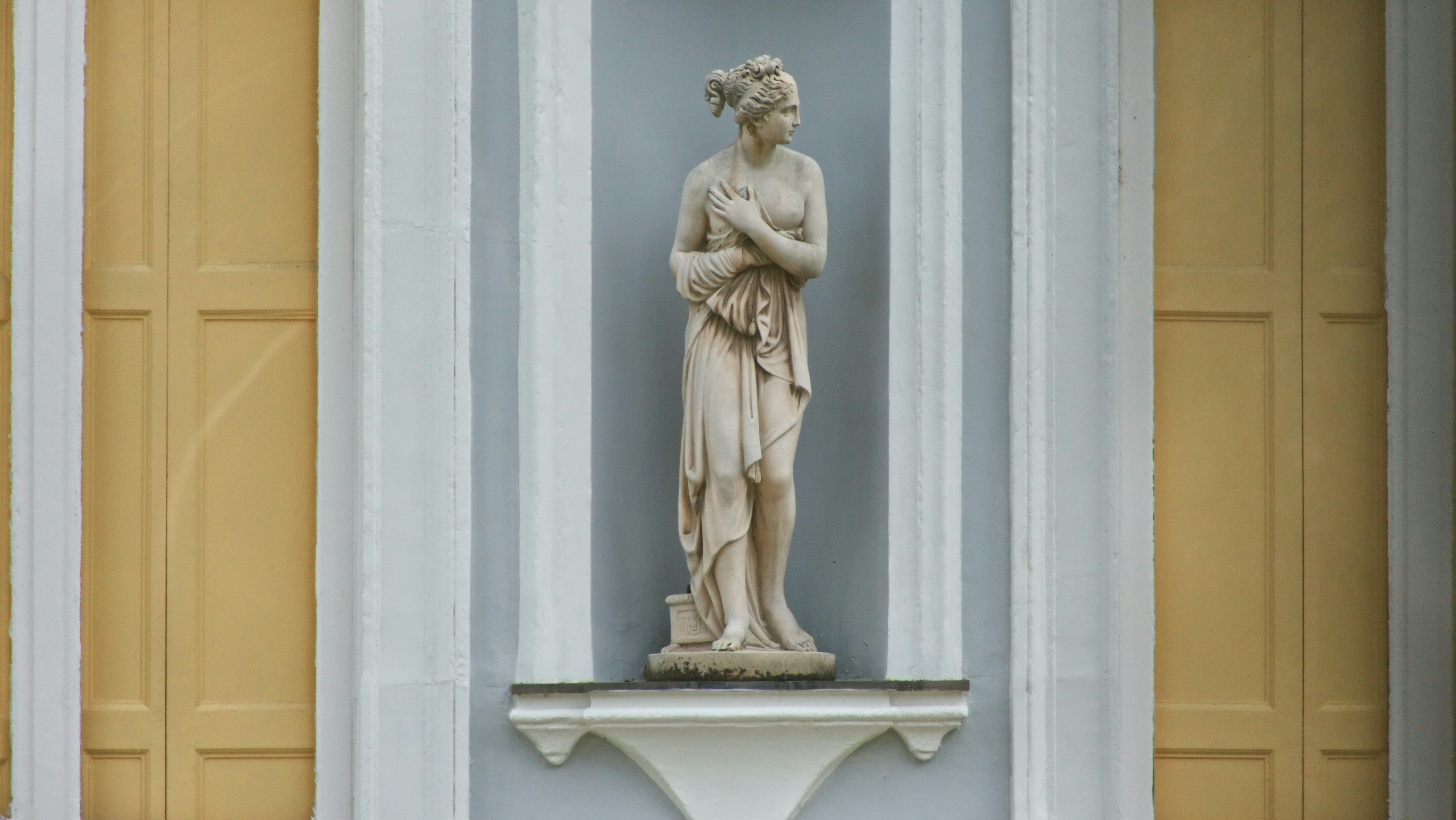 Classical statue of a woman in a niche, framed by contrasting yellow walls. The sculpture exhibits intricate details and a serene expression.