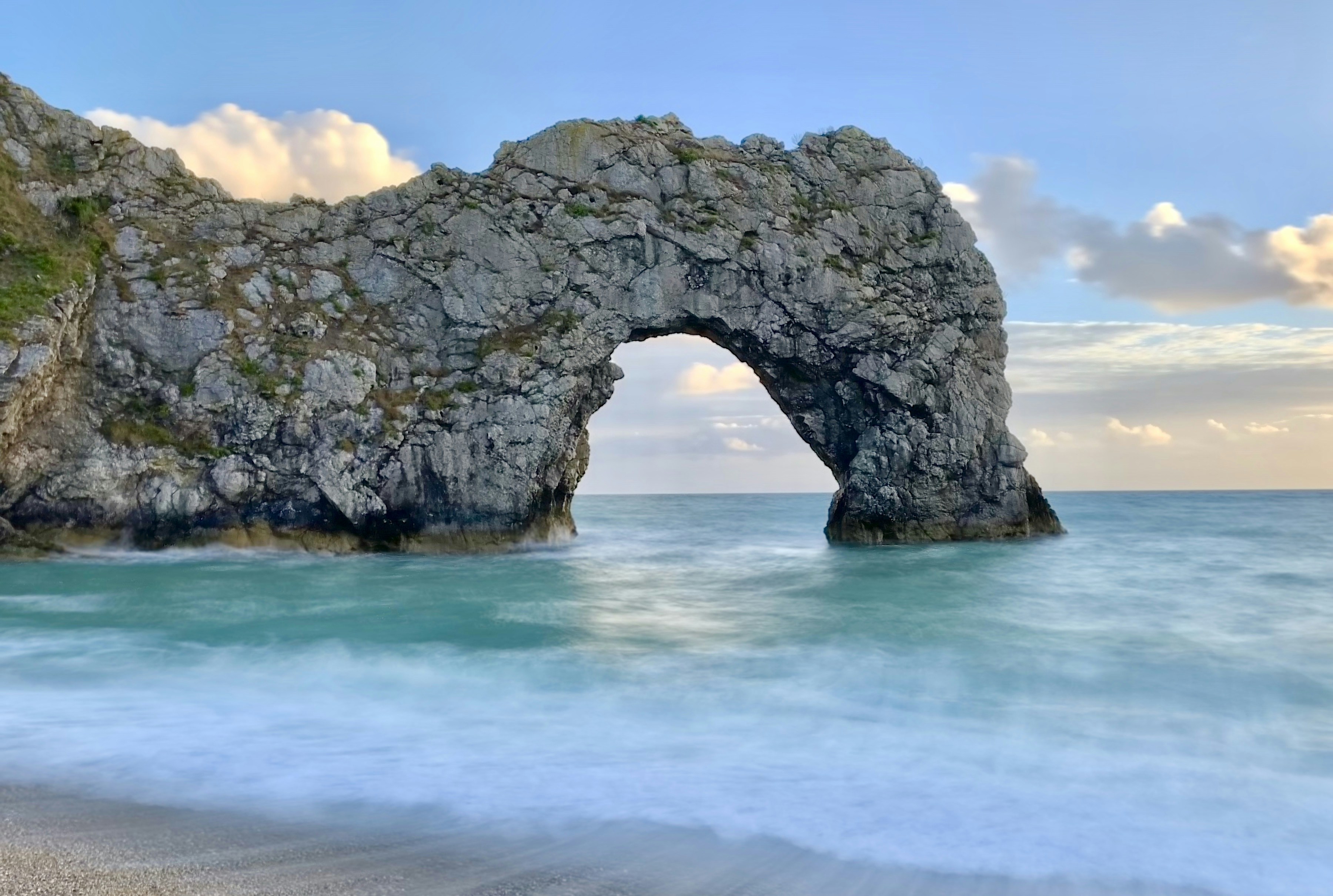 Rocky arch formation rising from the ocean, framed by gentle waves and a pastel sky at dusk.