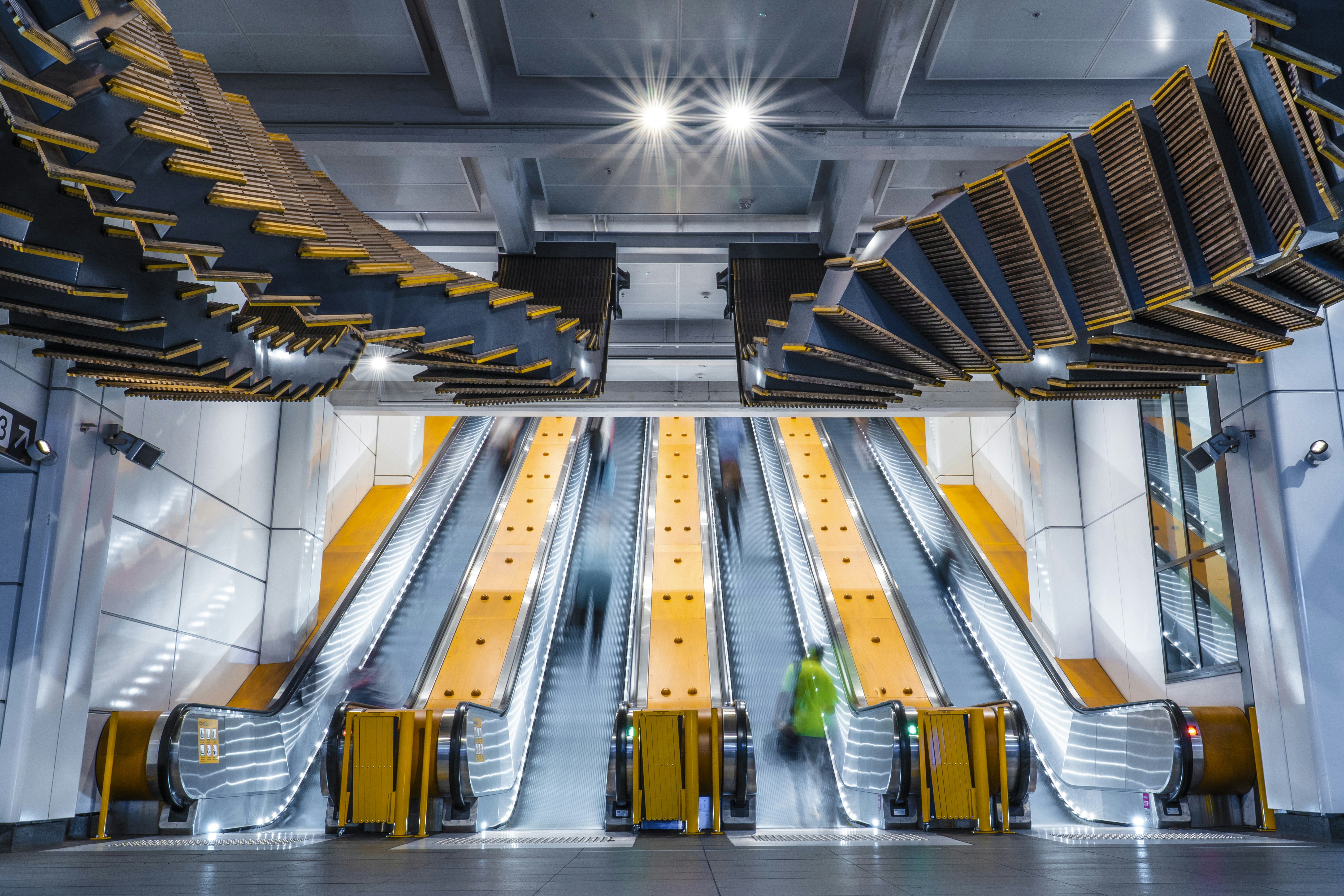 Vibrant escalators with blurred figures in a modern architectural space under geometric ceiling art.