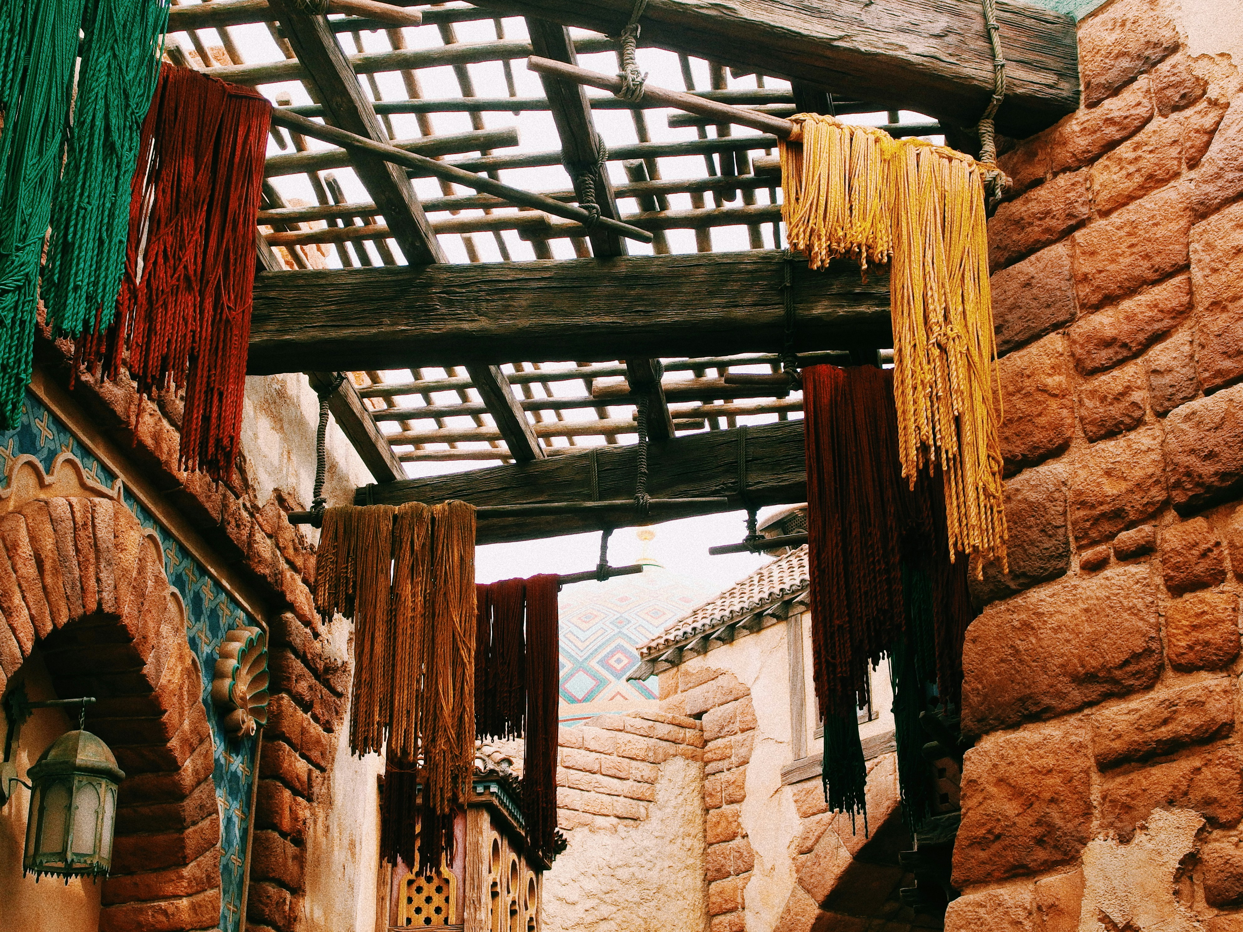 Colorful yarns hang between weathered brick walls and wooden beams in a sunlit alley, forming a vibrant corridor of texture and light. The scene highlights craft materials set against rustic architecture.