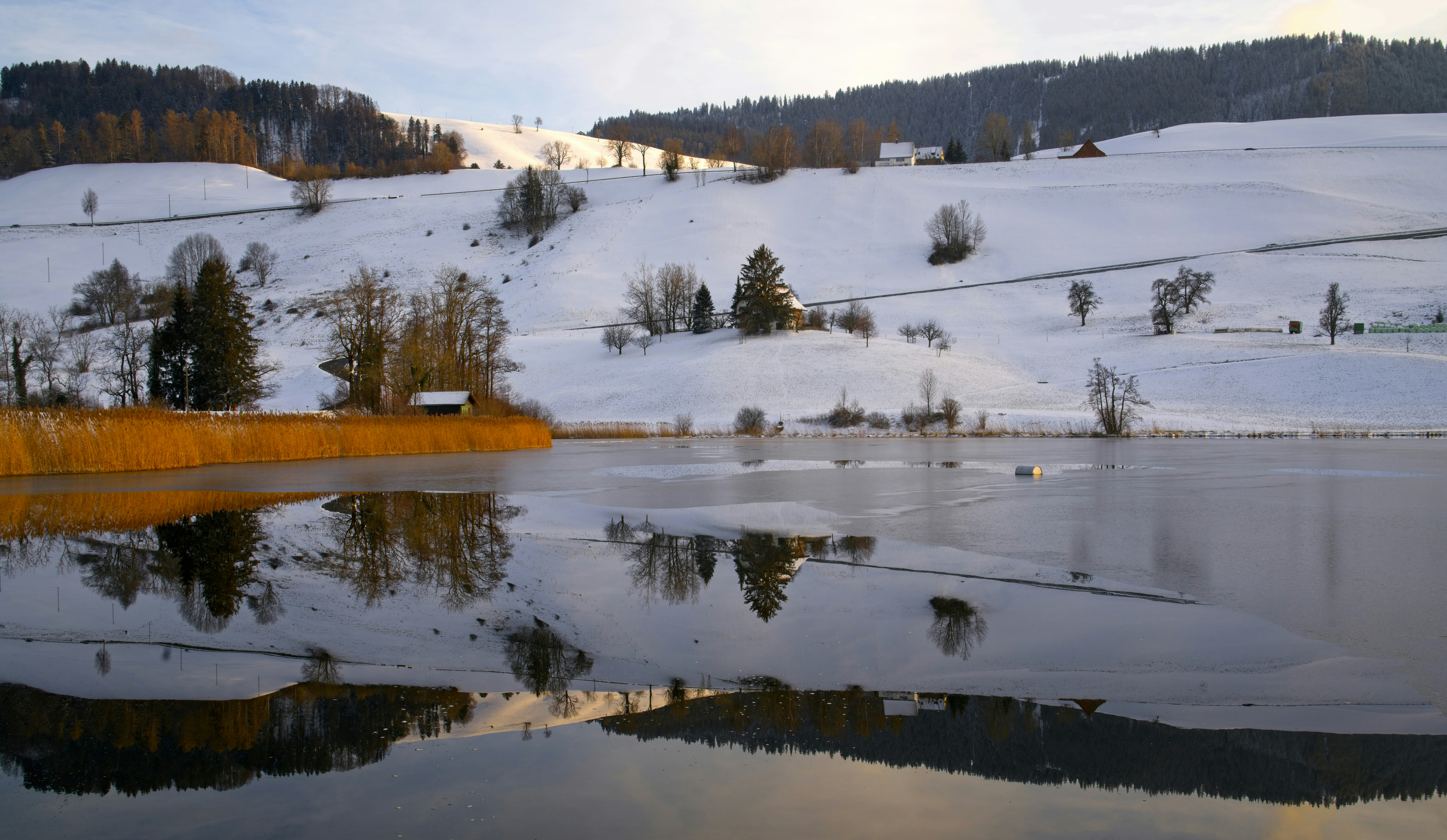 Snow-covered hills reflect in a calm, partially frozen lake under a serene sky.