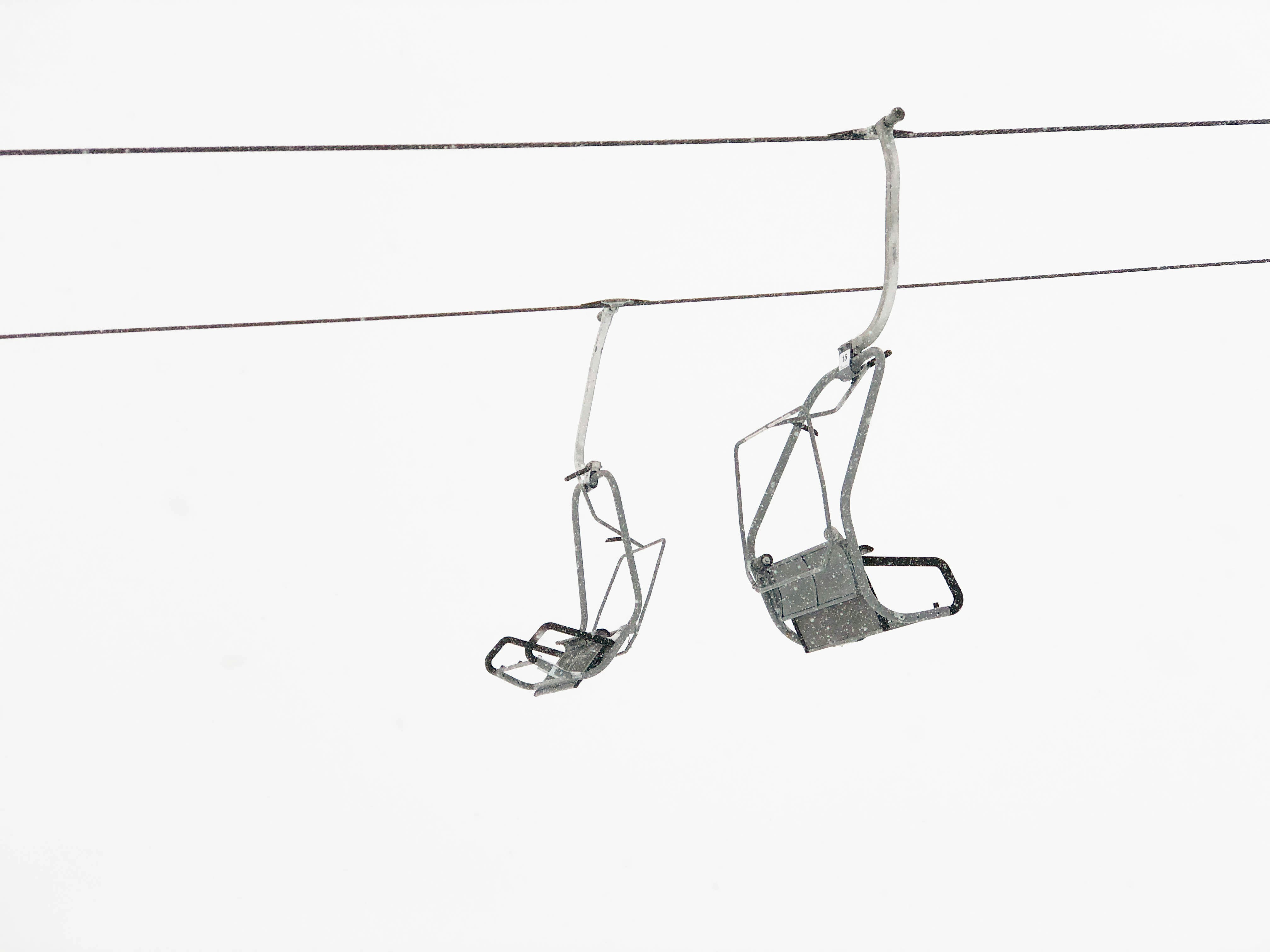 Two empty ski lift chairs suspended against a white, snowy backdrop, evoking a sense of stillness and solitude.