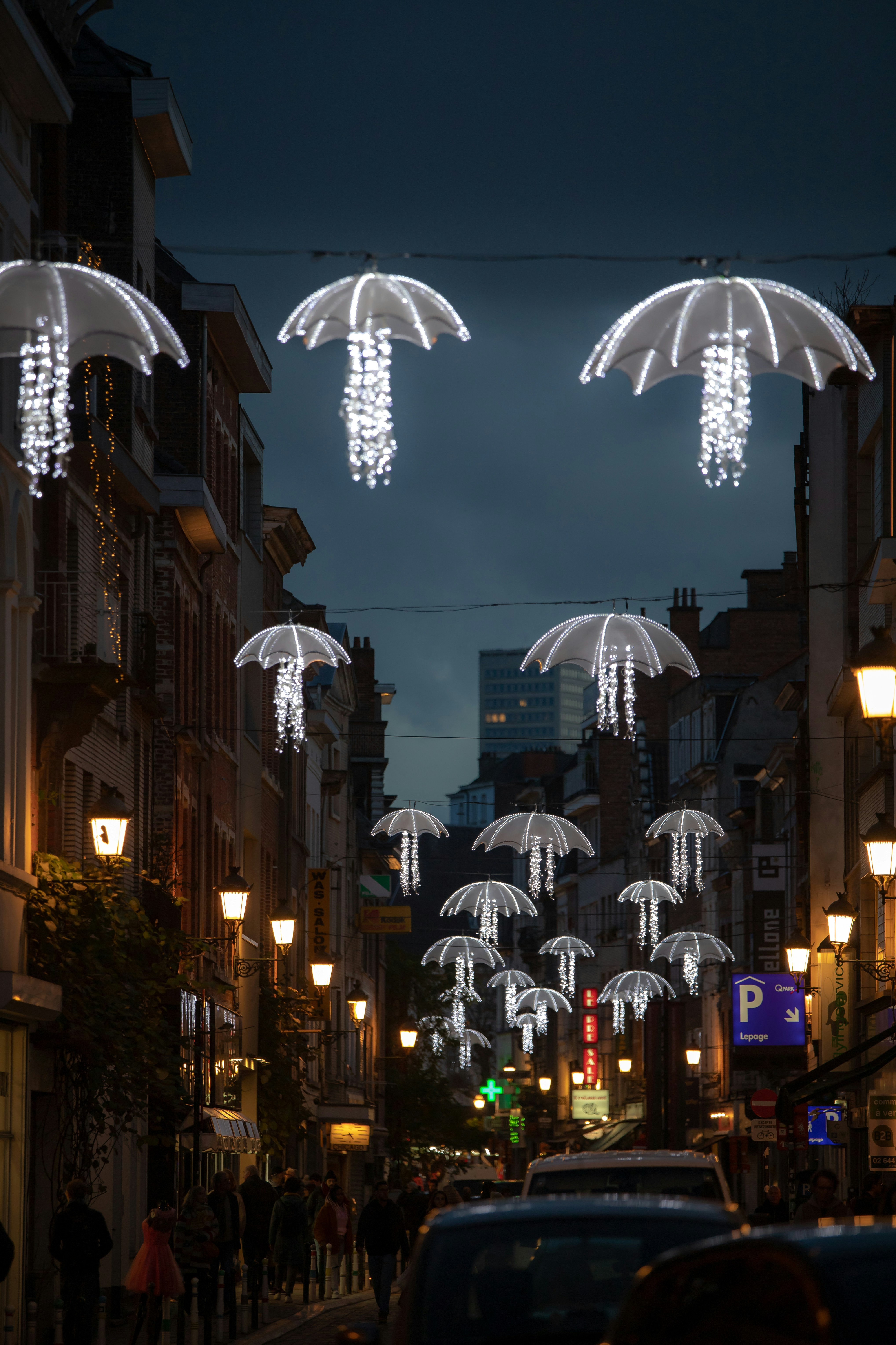 Festive umbrellas adorned with lights hang above a bustling street, creating a whimsical atmosphere during twilight. The scene captures the charm of urban nightlife.