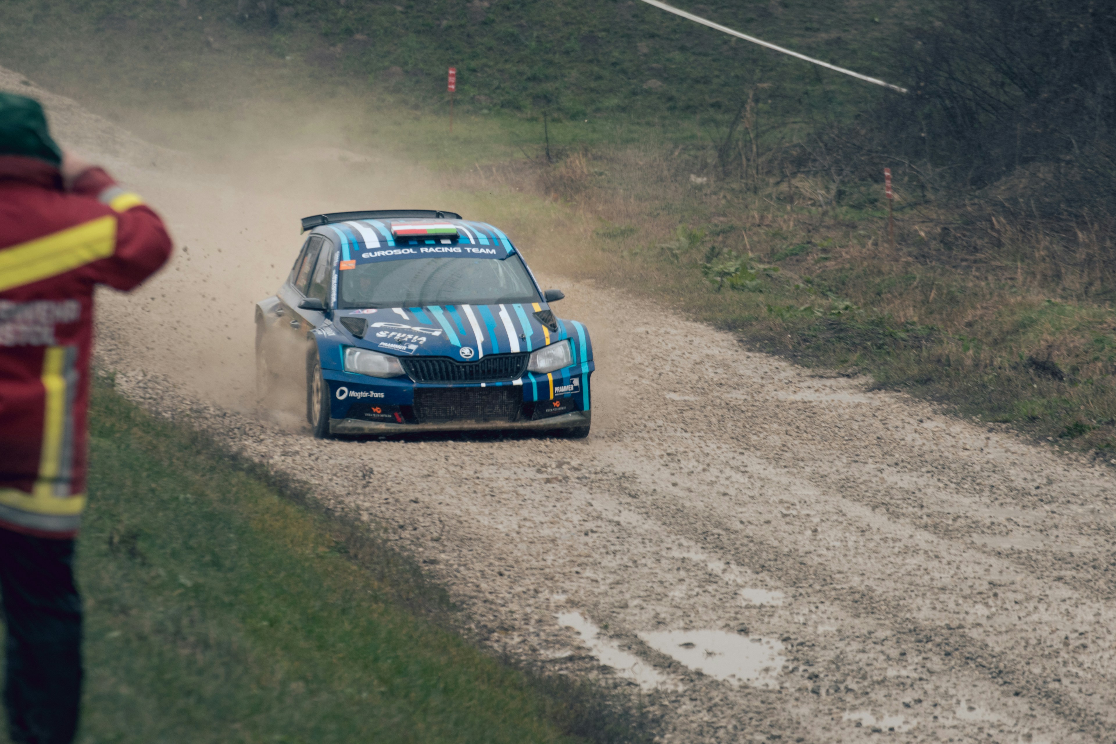 Blue rally car speeding on a gravel road with dust trailing behind.