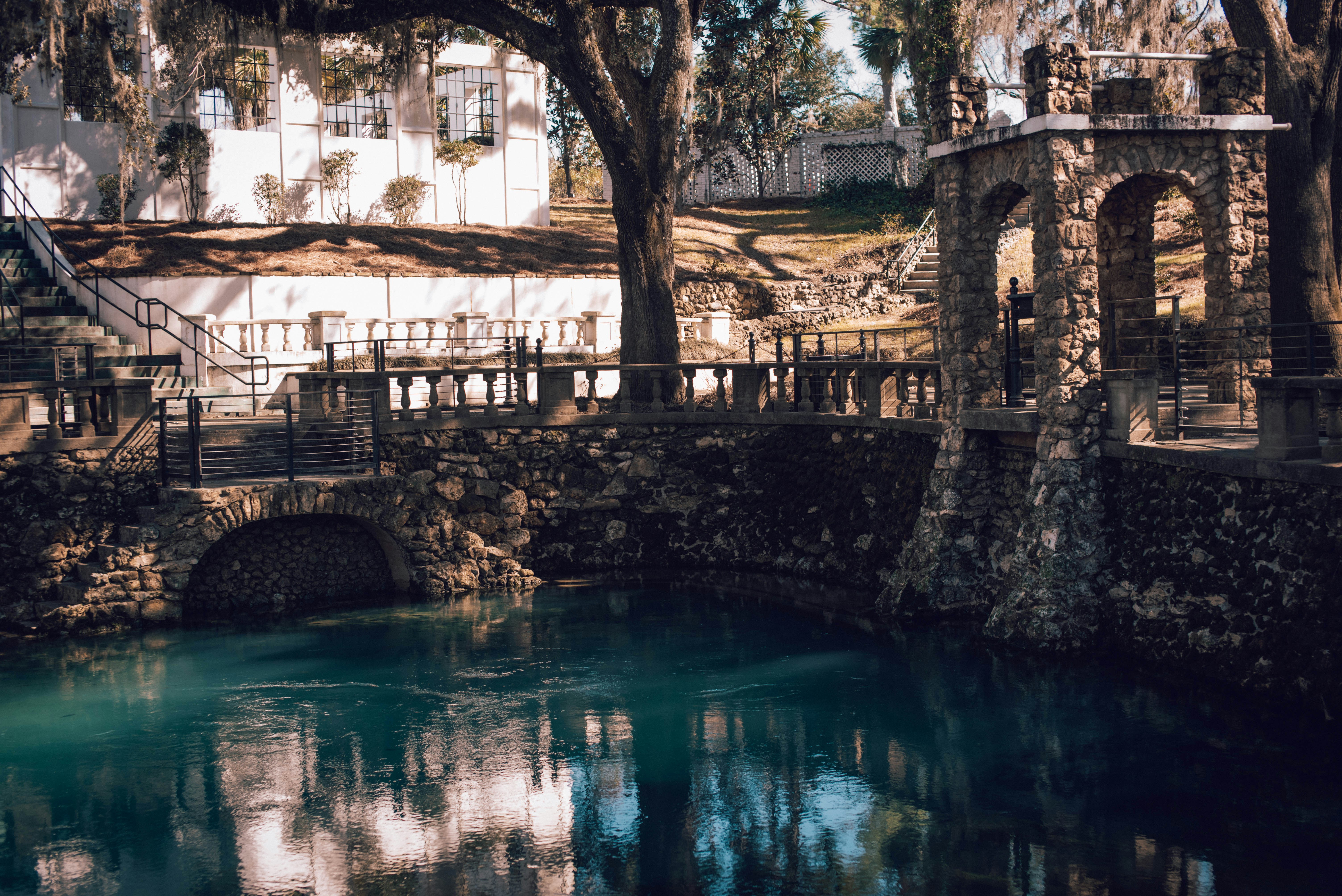 Stone bridge arches over tranquil azure waters surrounded by ancient trees.