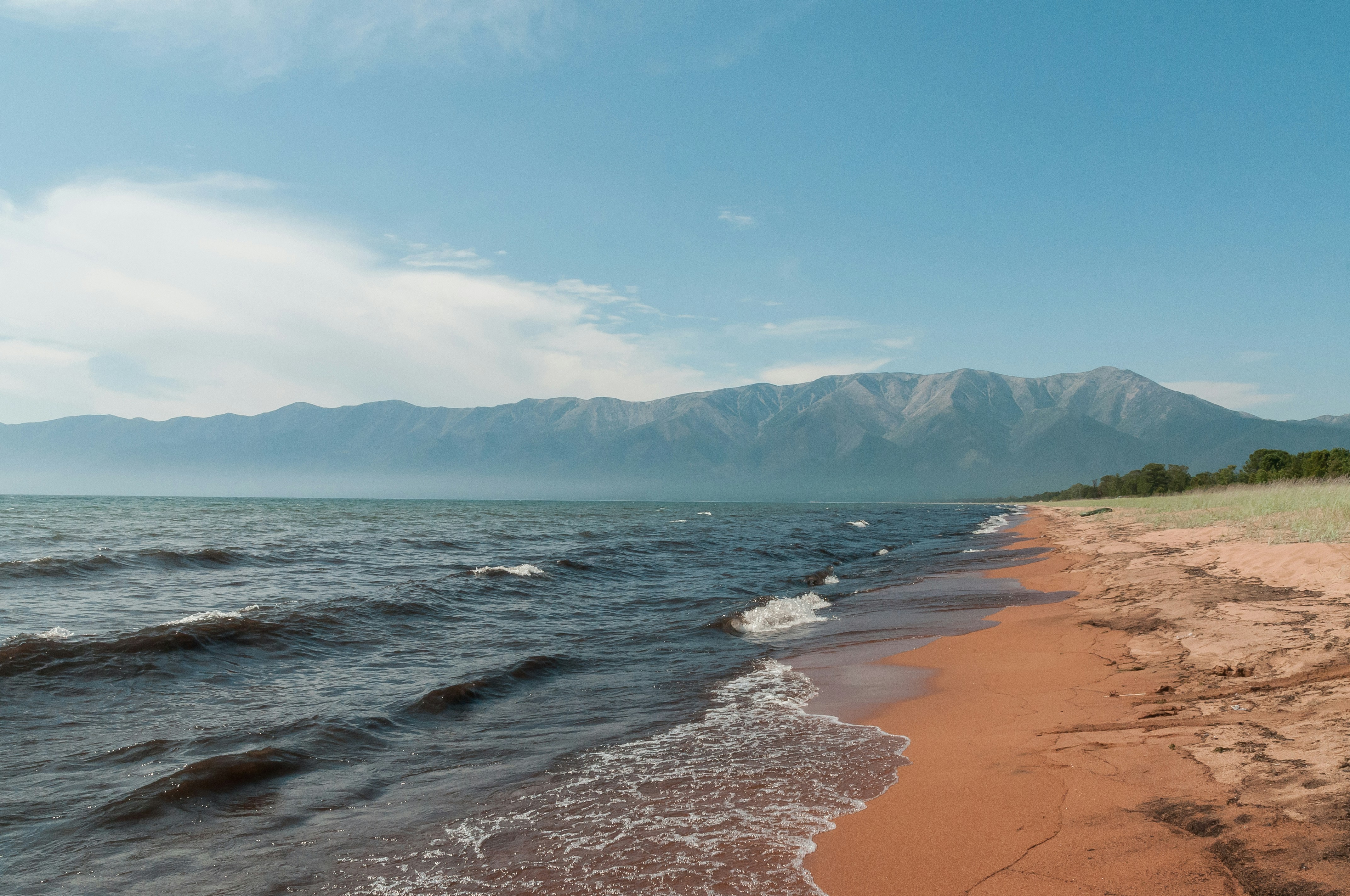 Sandy shoreline meeting gentle waves under a clear sky with distant mountains.