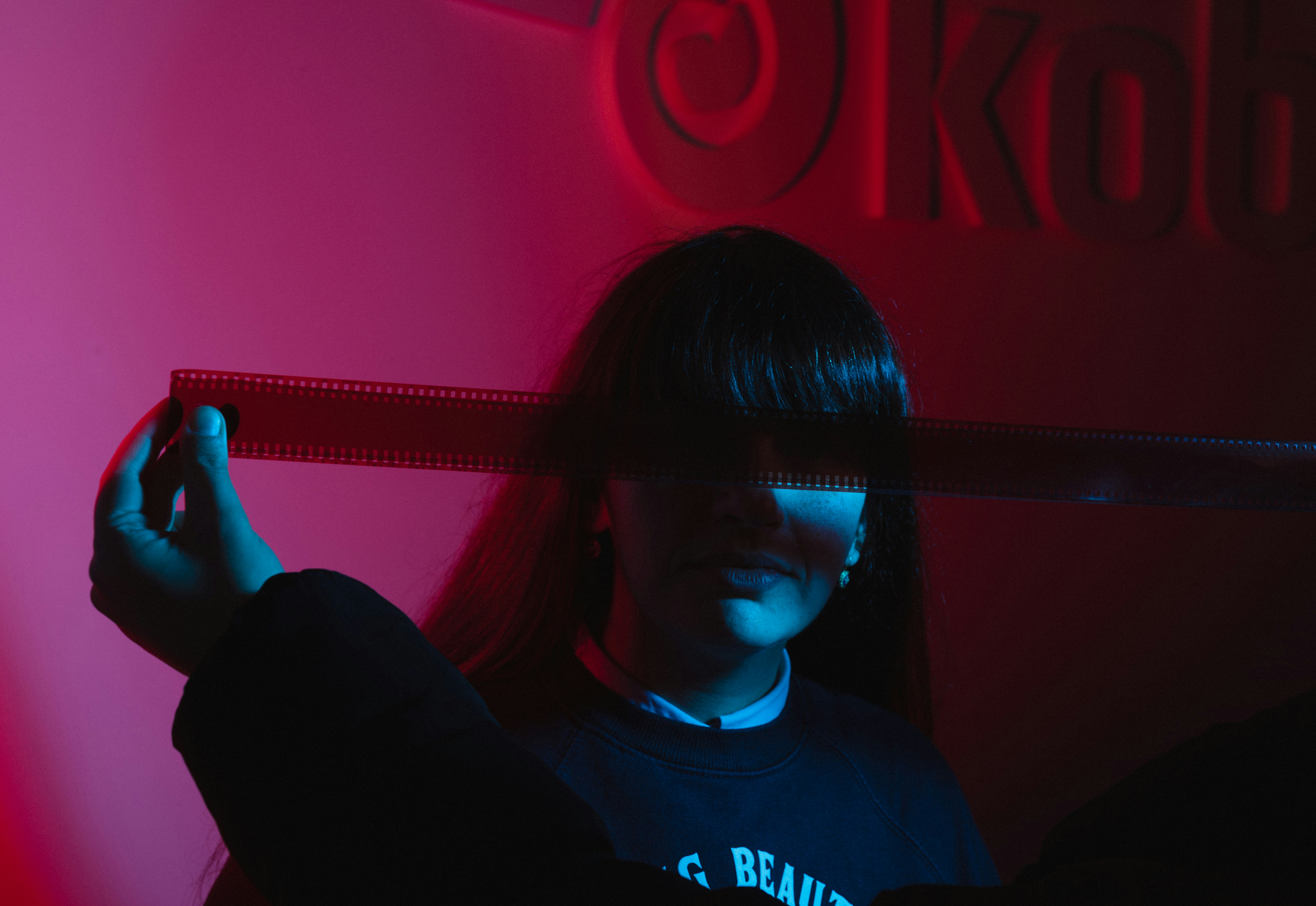 A young person holds a film strip in front of their face, illuminated by vibrant red and blue lighting, creating a striking contrast against a softly blurred background.