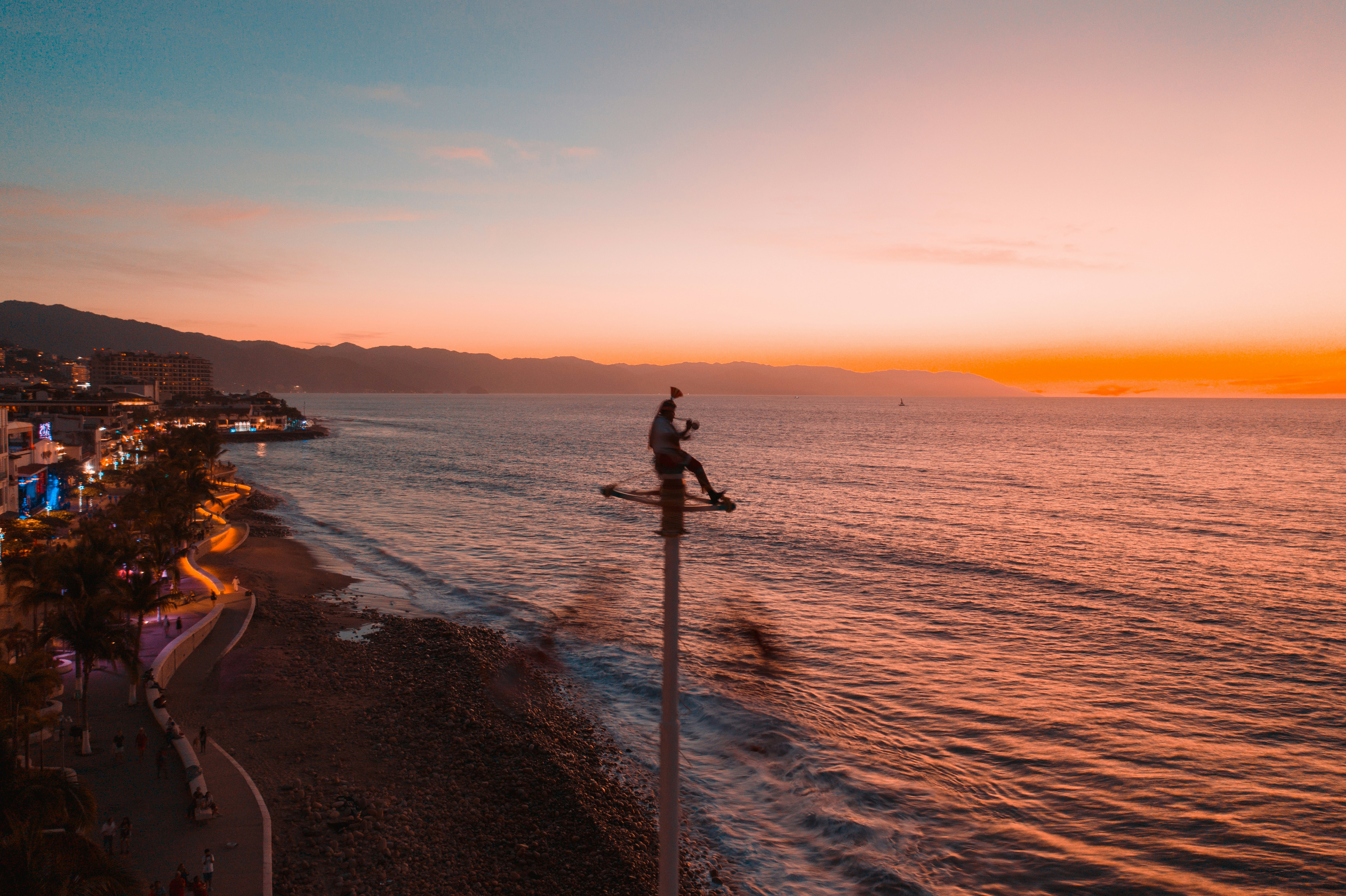 Performers in traditional attire descend from a tall pole over a coastal landscape at sunset.