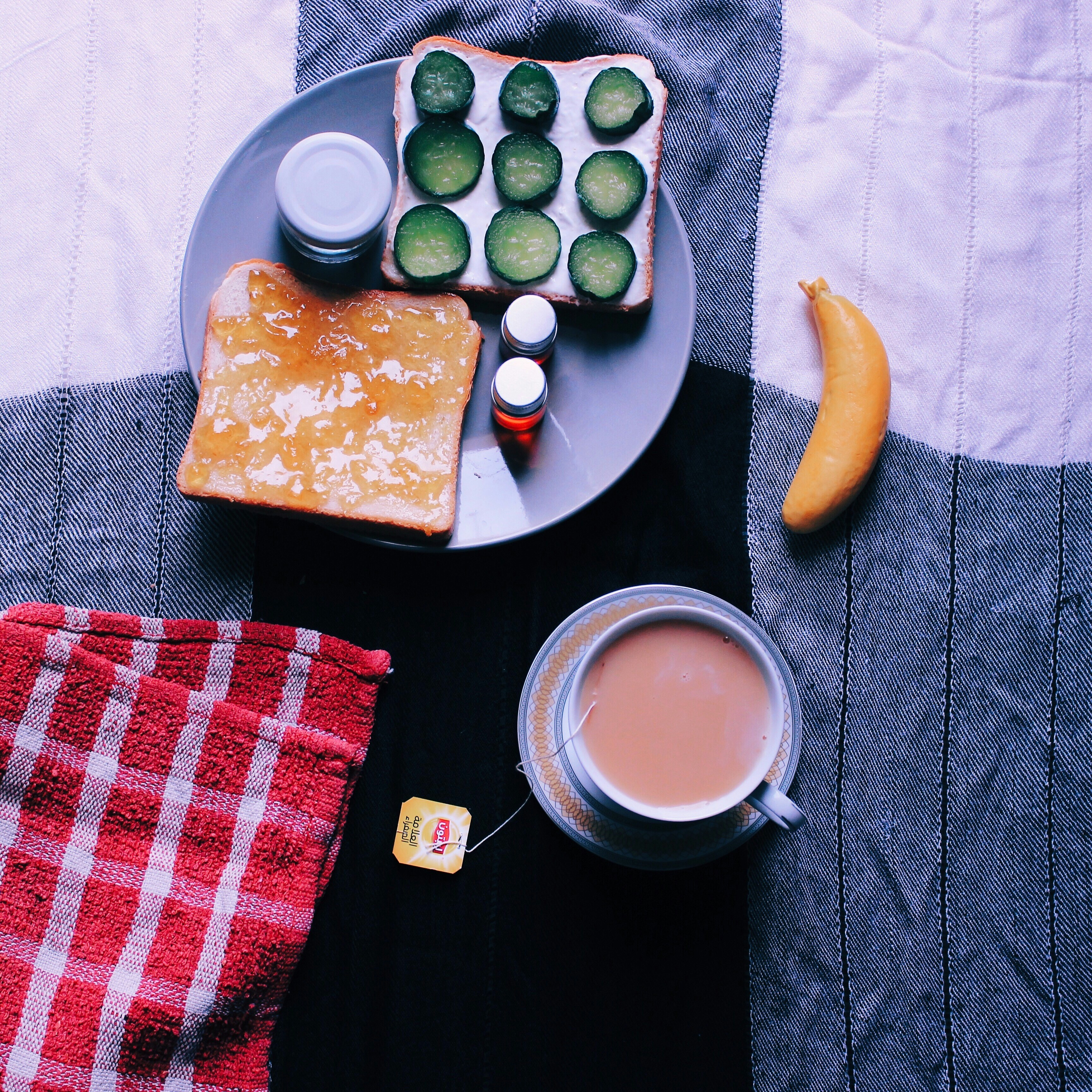 A cozy breakfast scene featuring a plate of cucumber-topped toast, a jar of jam, colorful vitamins, a banana, and a cup of tea, all set against a patterned blanket.