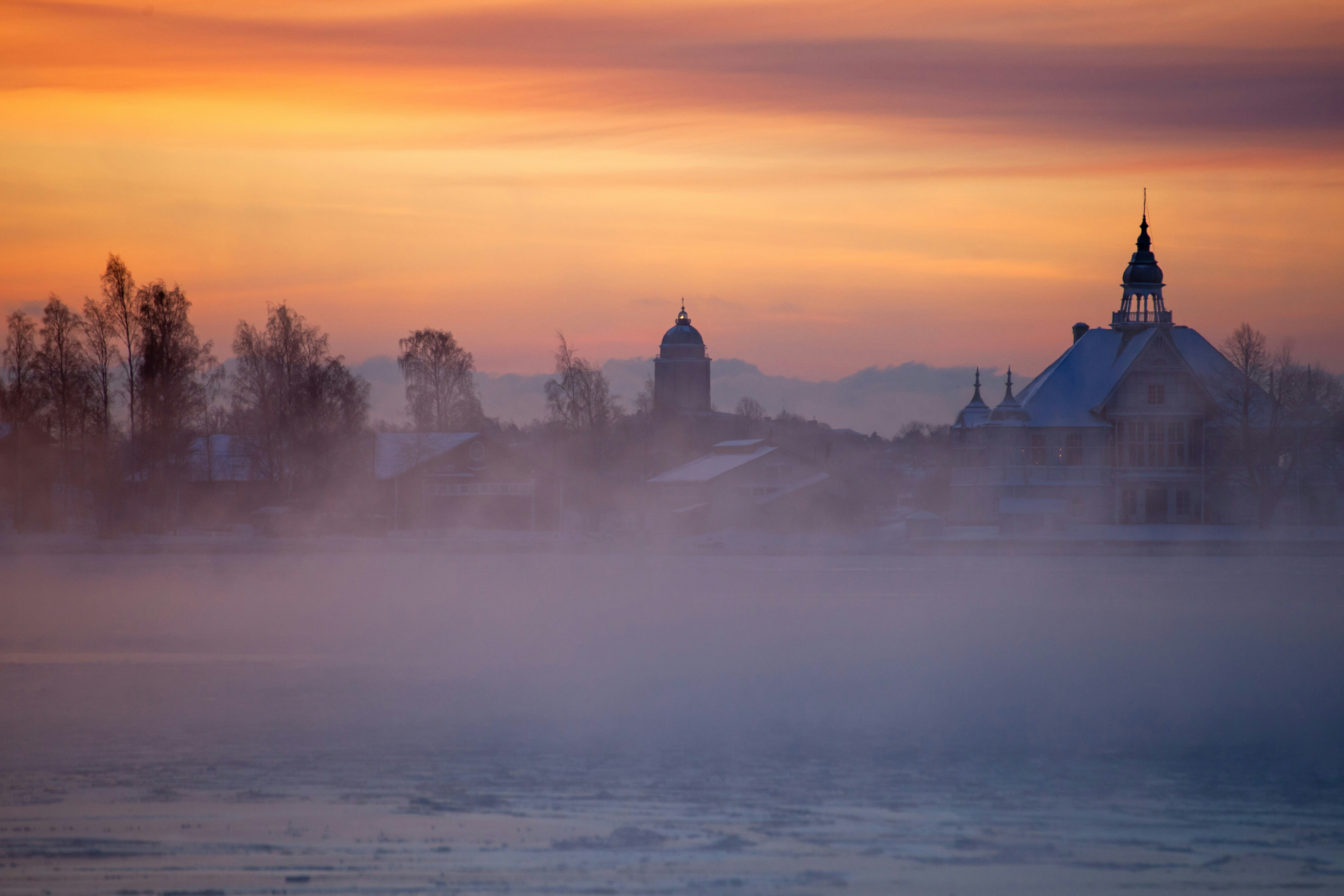 Misty winter landscape with a silhouette of a historic building against a vibrant sunset sky. The ethereal fog blankets the water, creating a serene atmosphere.