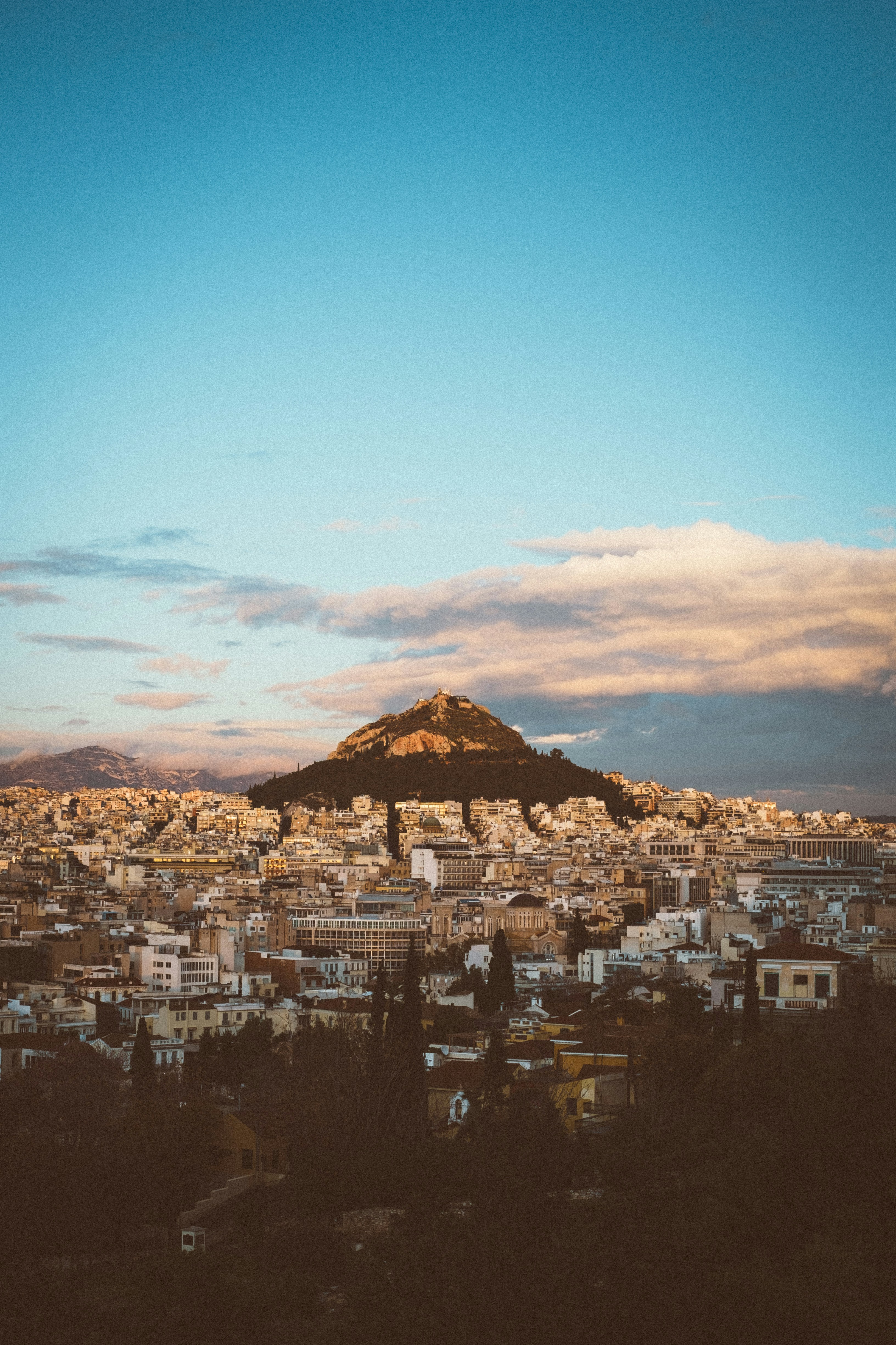 A panoramic view of Athens with Mount Lycabettus standing tall amidst the urban landscape, bathed in the warm glow of sunset.