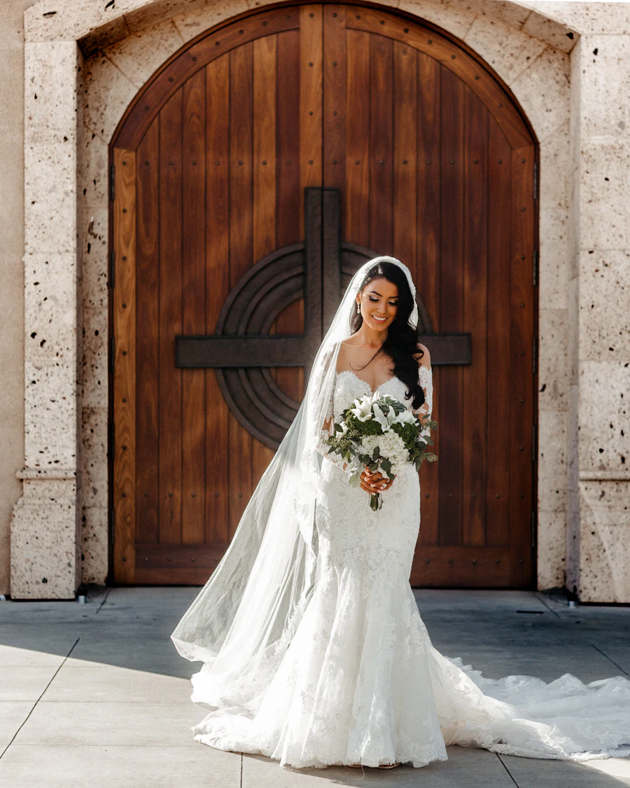 Bride in a white lace gown and veil stands smiling with a bouquet in front of large arched wooden doors.