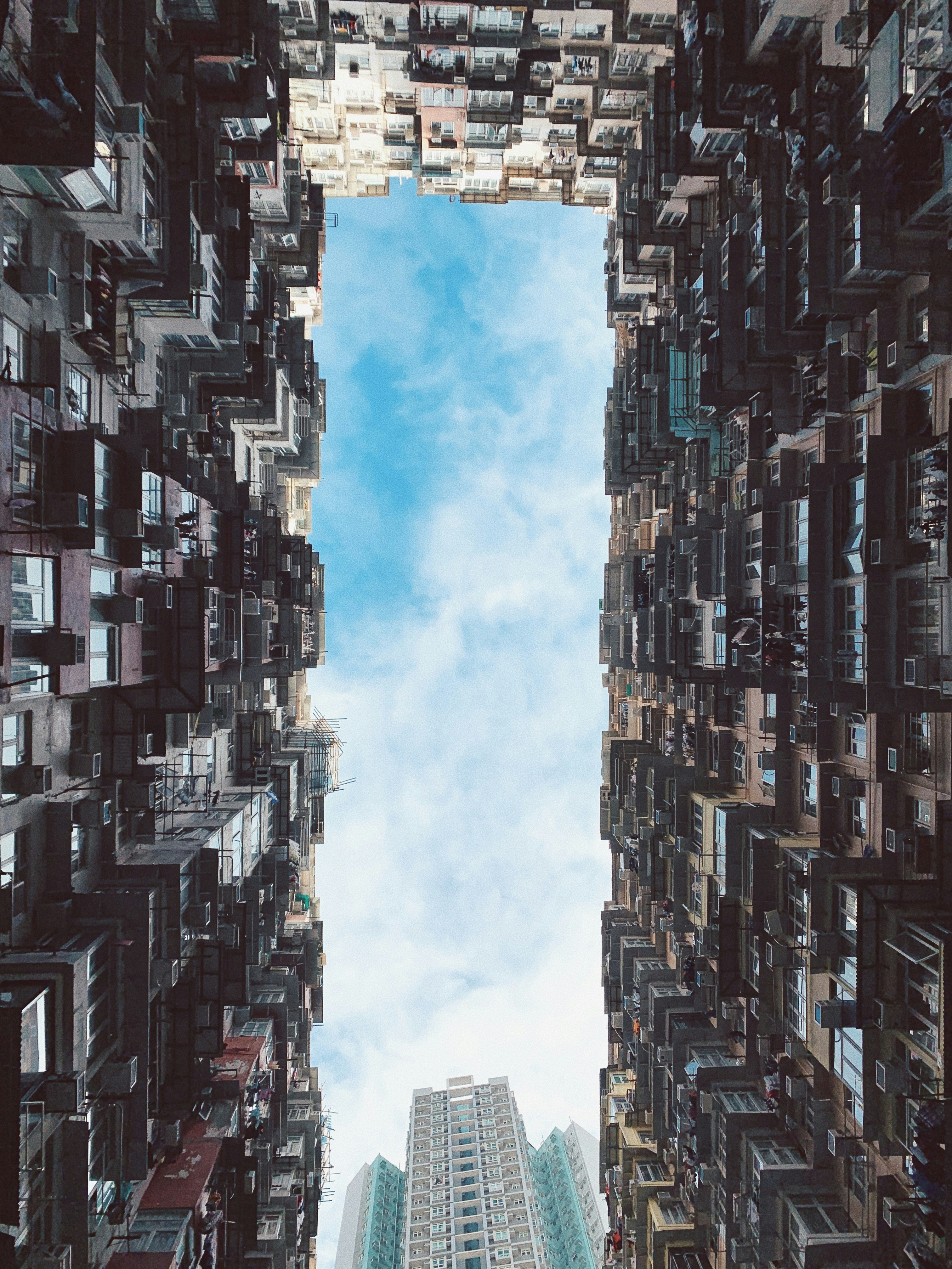 View looking up through a narrow urban canyon formed by densely packed residential buildings, revealing a patch of blue sky above.