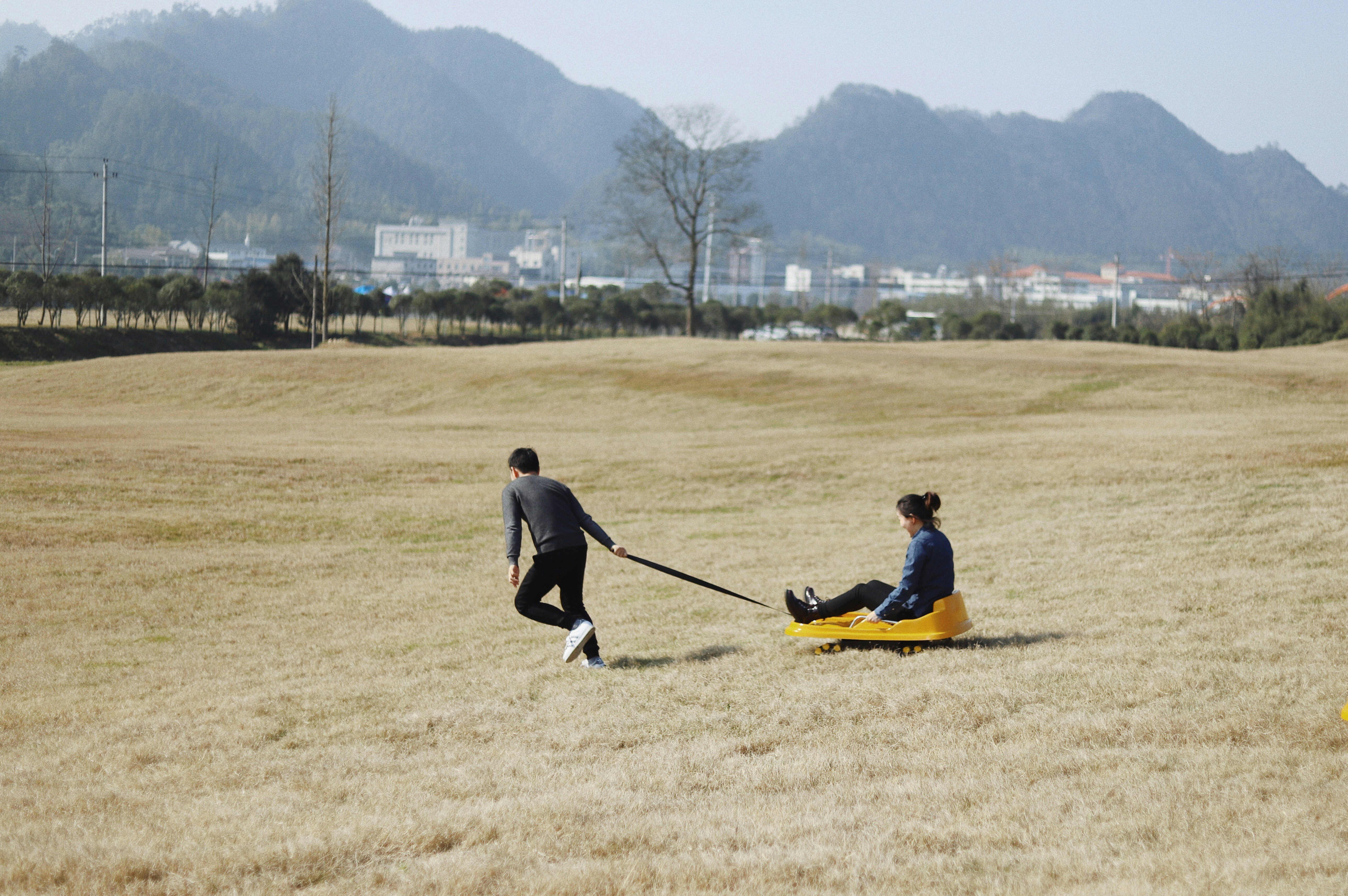 Two individuals enjoying a playful moment on a grassy field, with one pulling the other on a sled. The backdrop features rolling mountains and a clear sky.