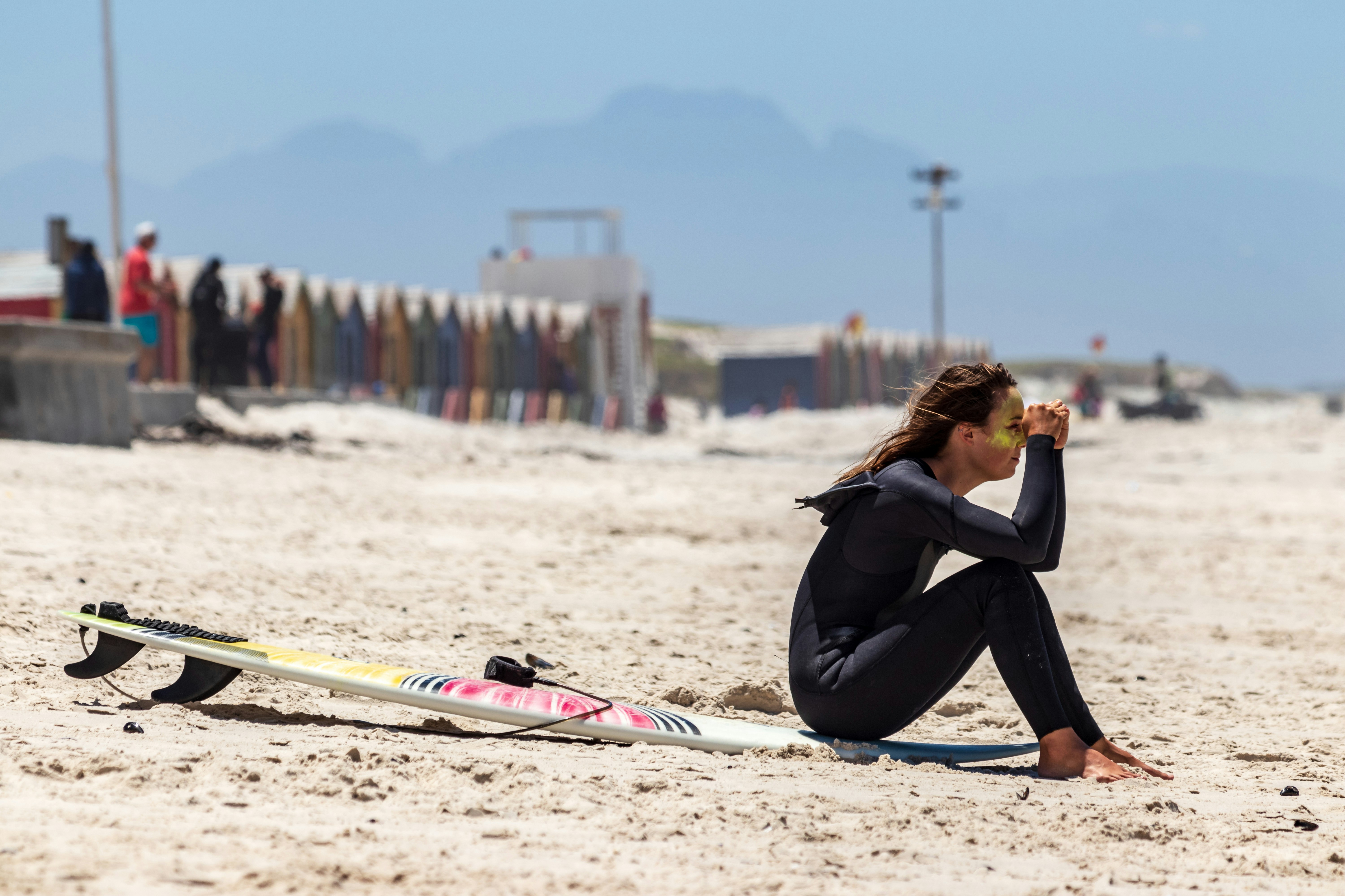 Surfer in a wetsuit sits pensively on the beach, with a surfboard beside her and colorful boards lined up in the background. The distant mountains add depth to the coastal scene.