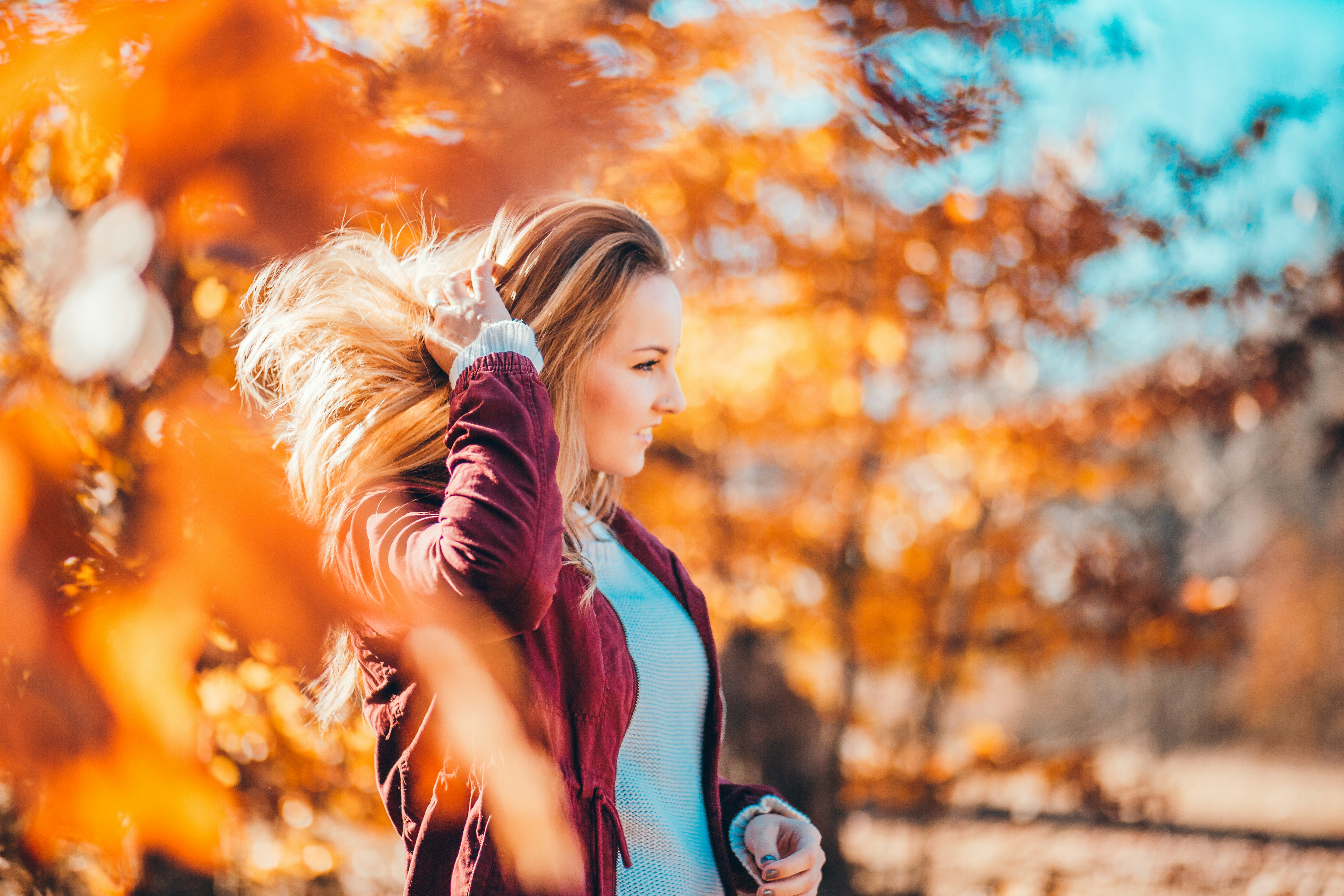Woman in a maroon jacket stands amidst vibrant autumn foliage, holding her hair against a bright blue sky.