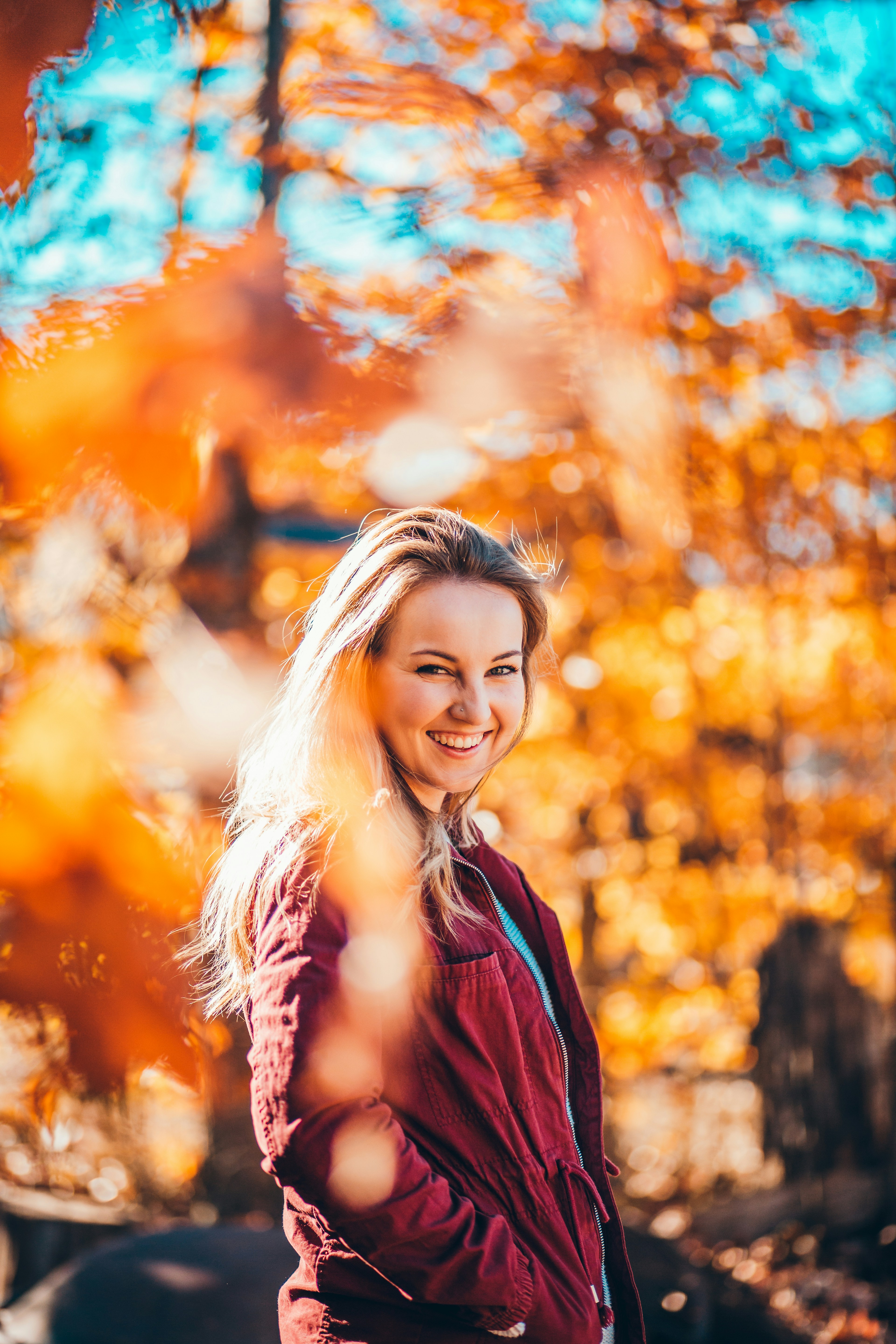 A smiling woman stands amidst vibrant autumn foliage, her joyful expression framed by golden leaves. The scene captures the warmth and beauty of fall.
