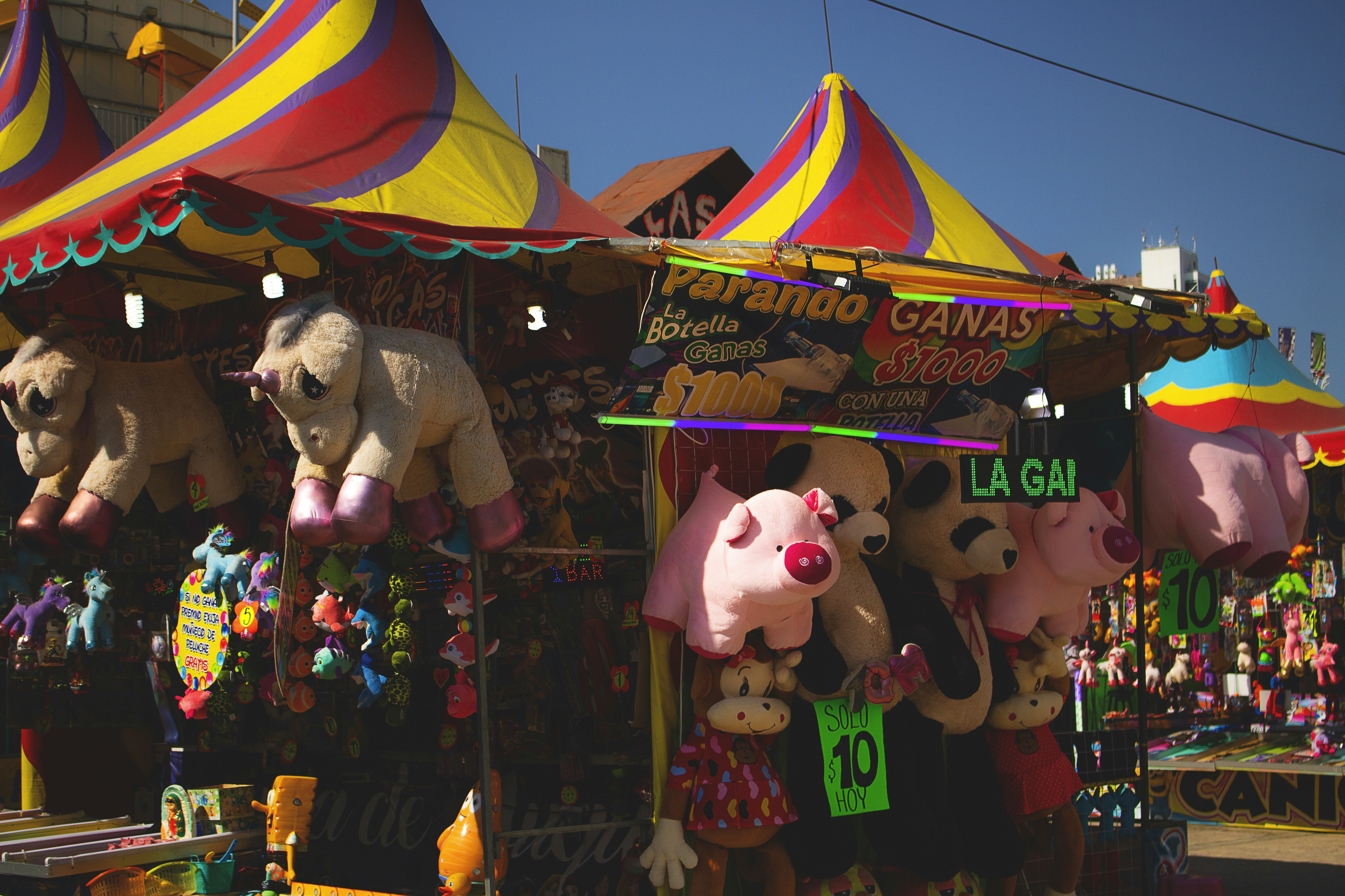 Carnival stall selling plush toys under striped canopies, with a pink pig centerpiece and bright signs.