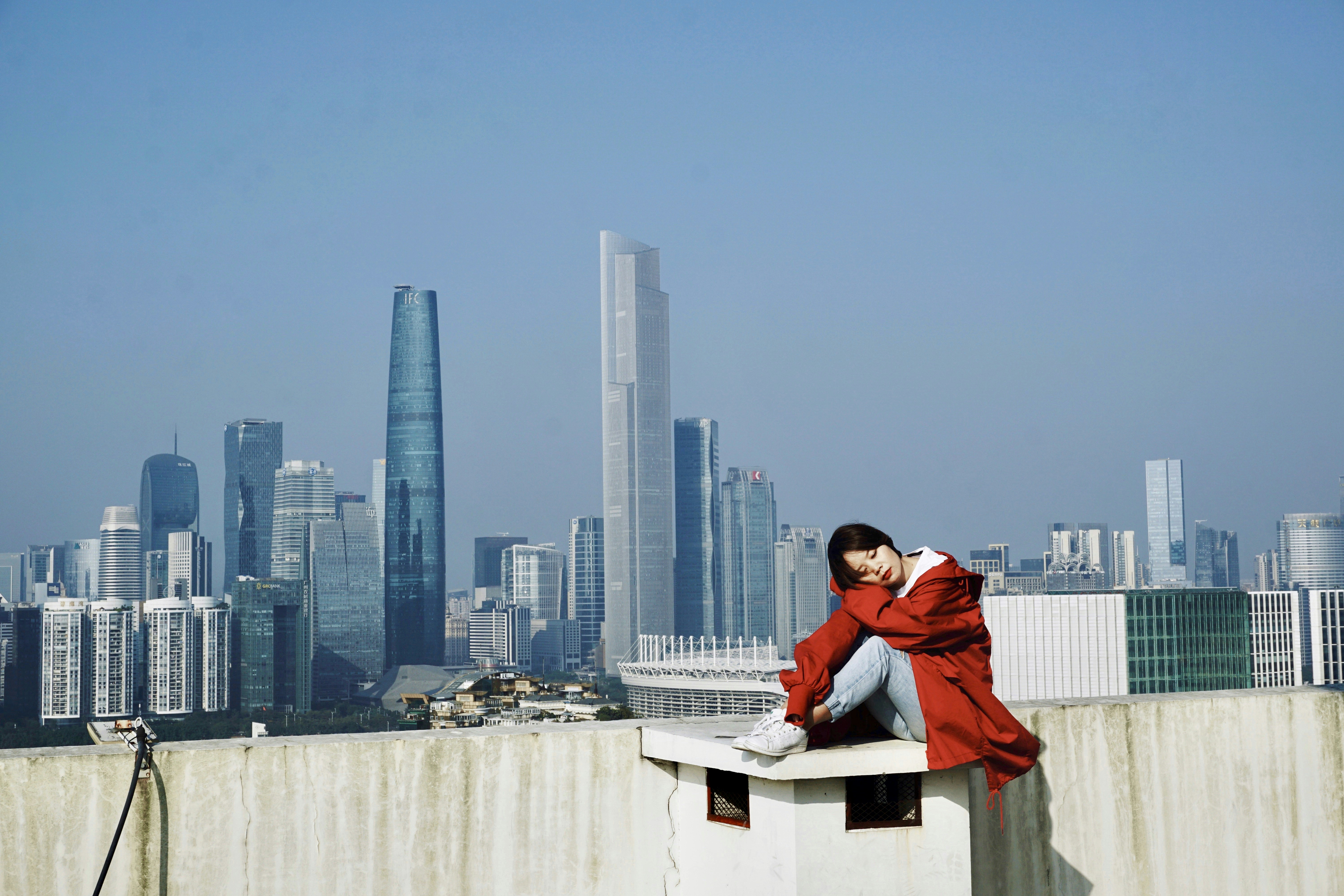 Person in red jacket sitting on a rooftop with a city skyline in the background.