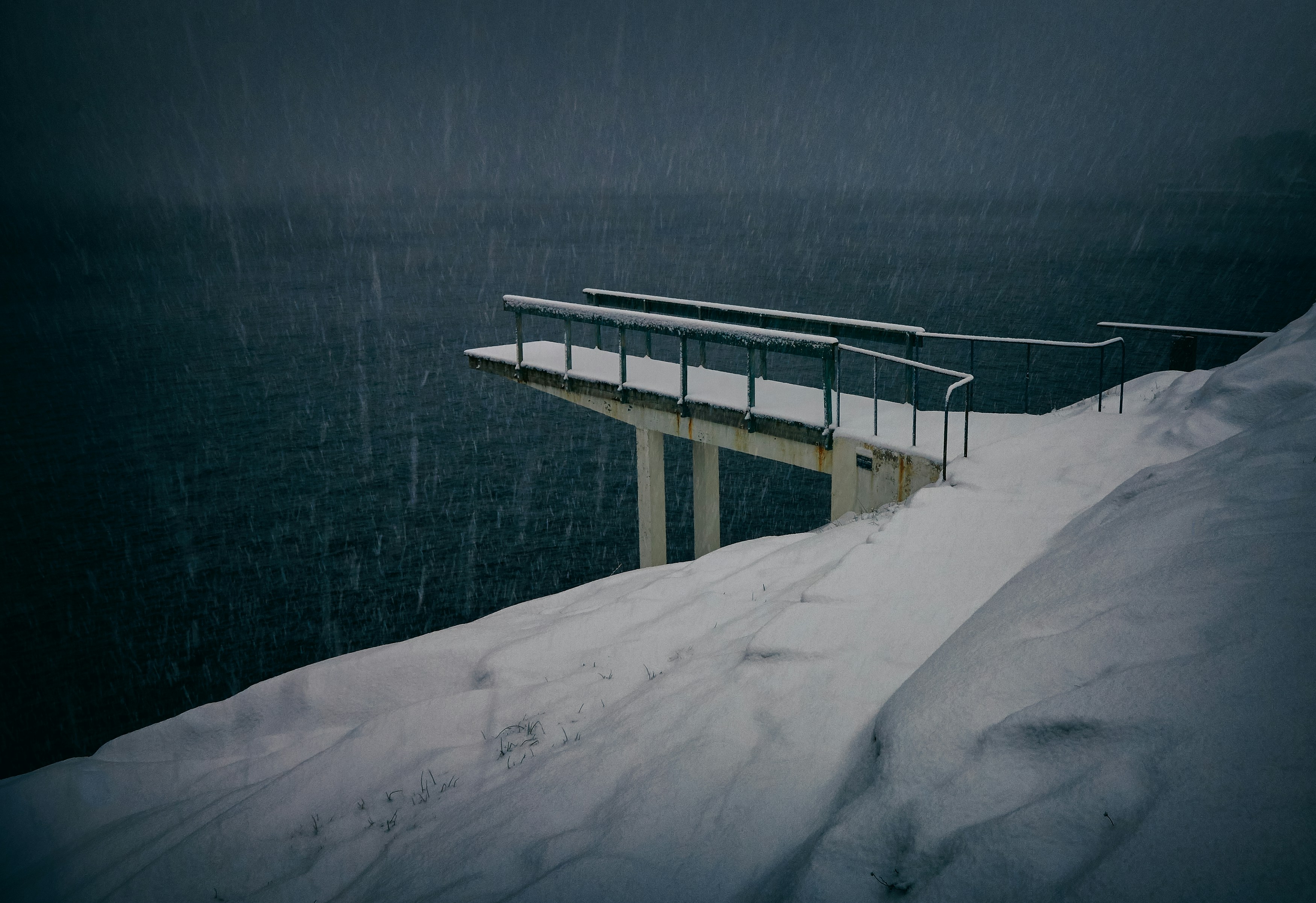 Snow-covered dock extending into a dark, icy lake under a heavy snowfall. The scene evokes a sense of solitude and stillness.