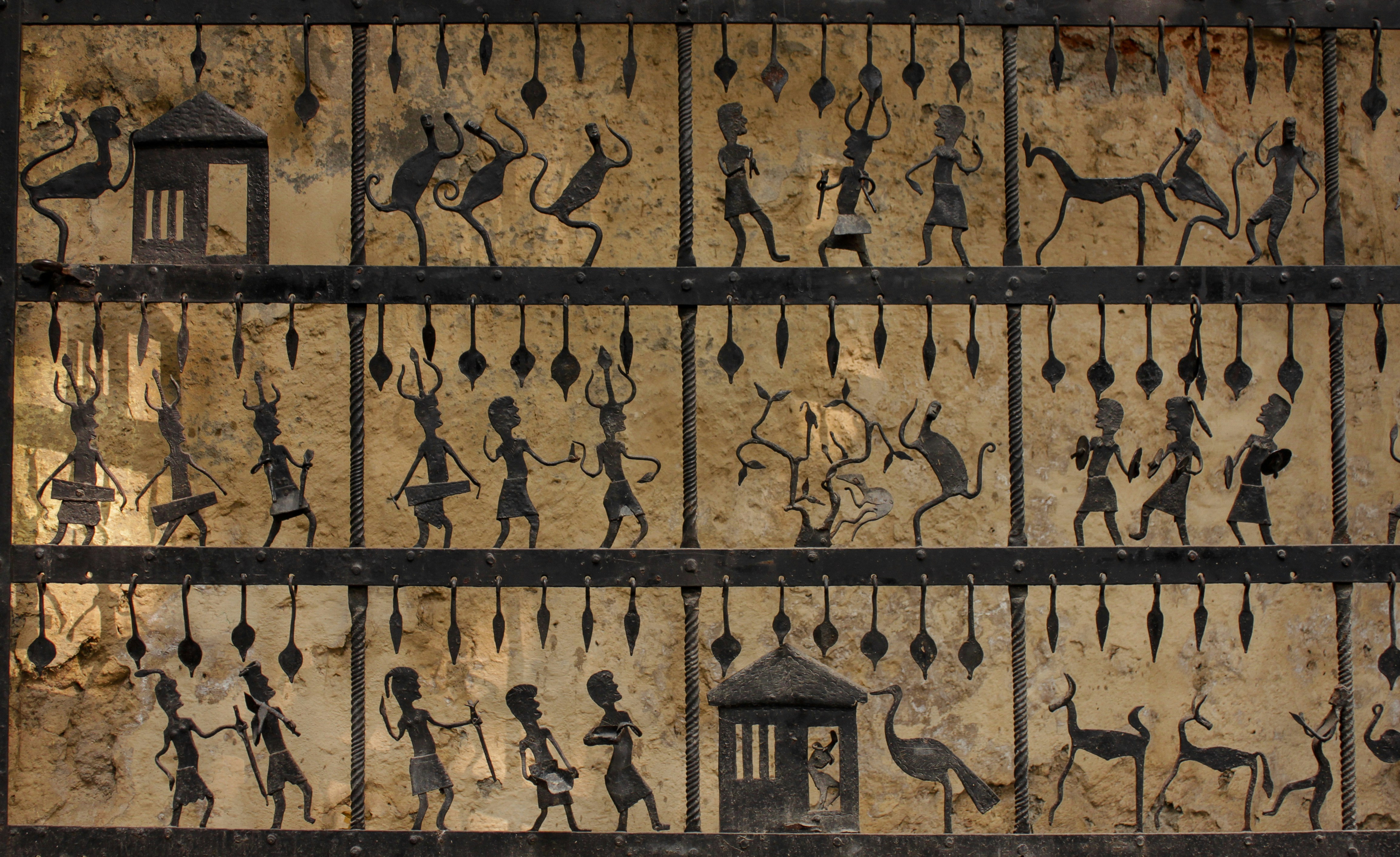 Intricate black silhouettes depicting traditional life and rituals, arranged in a rhythmic pattern on an aged wall. The artwork conveys cultural stories through its detailed figures.
