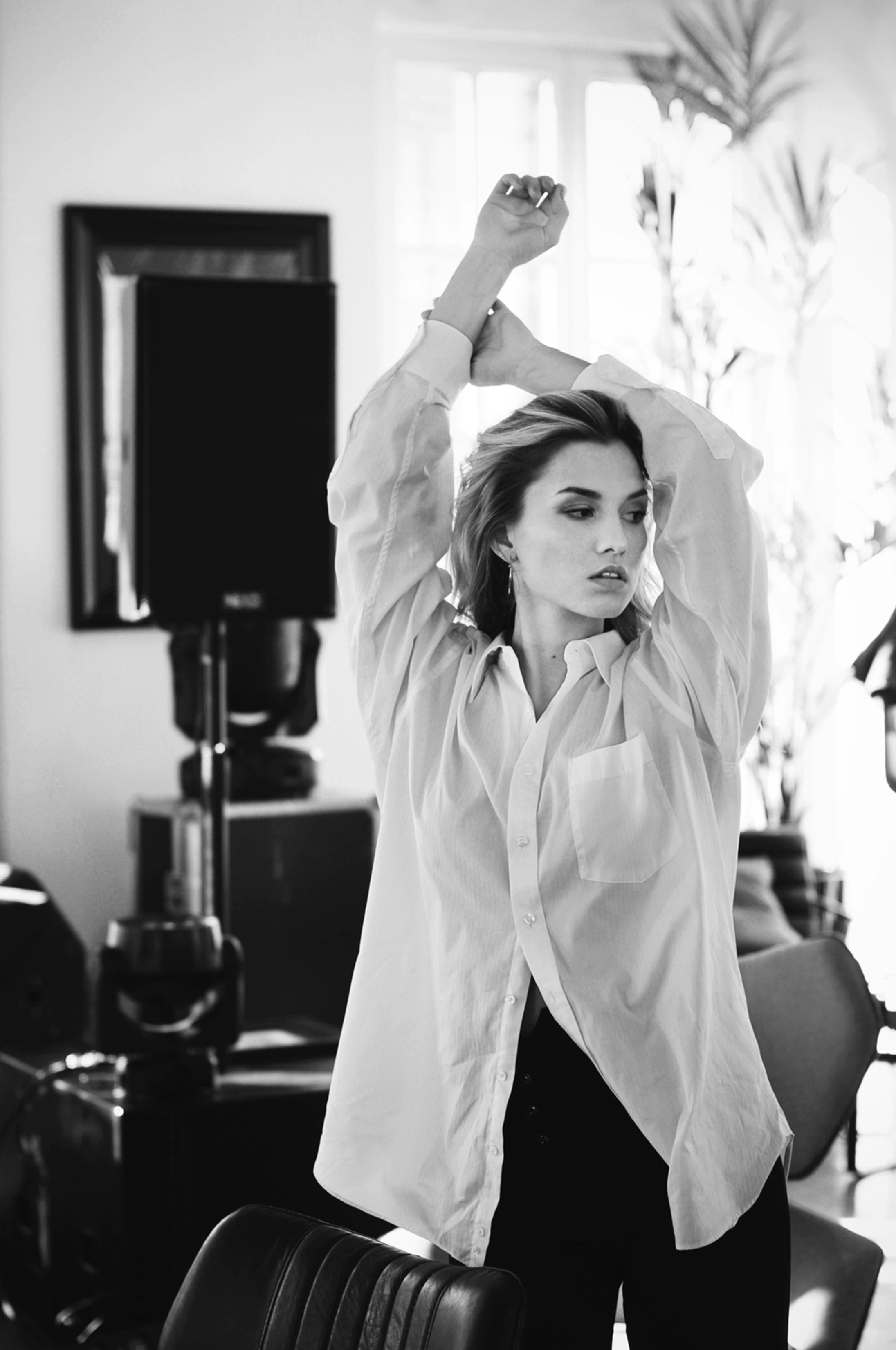 A woman in a white shirt poses with her arms raised, exuding a sense of grace and confidence in a stylish interior setting.