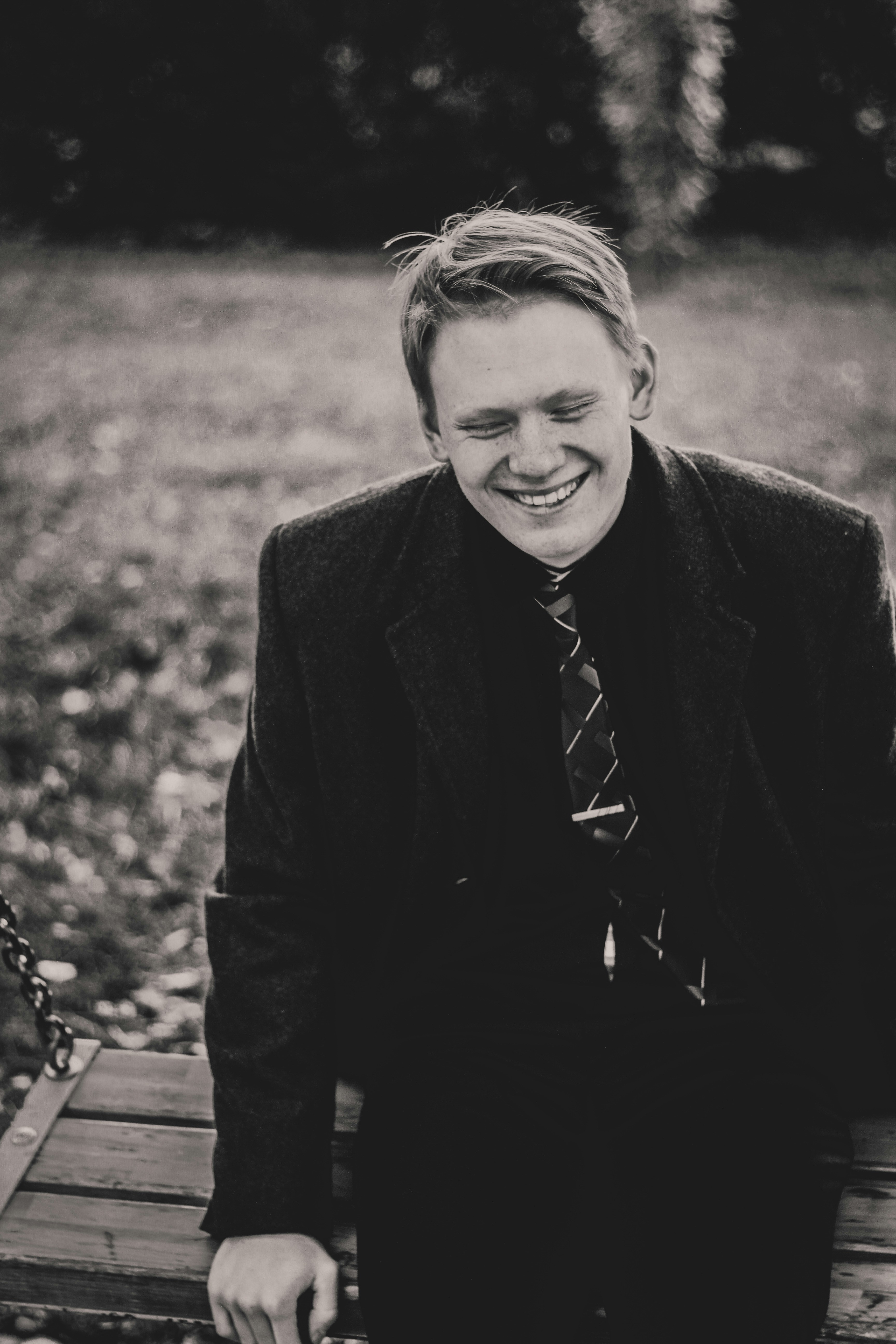 Smiling young man seated on a wooden bench, dressed in formal attire with a playful expression. The image captures a moment of happiness amidst a serene outdoor setting.