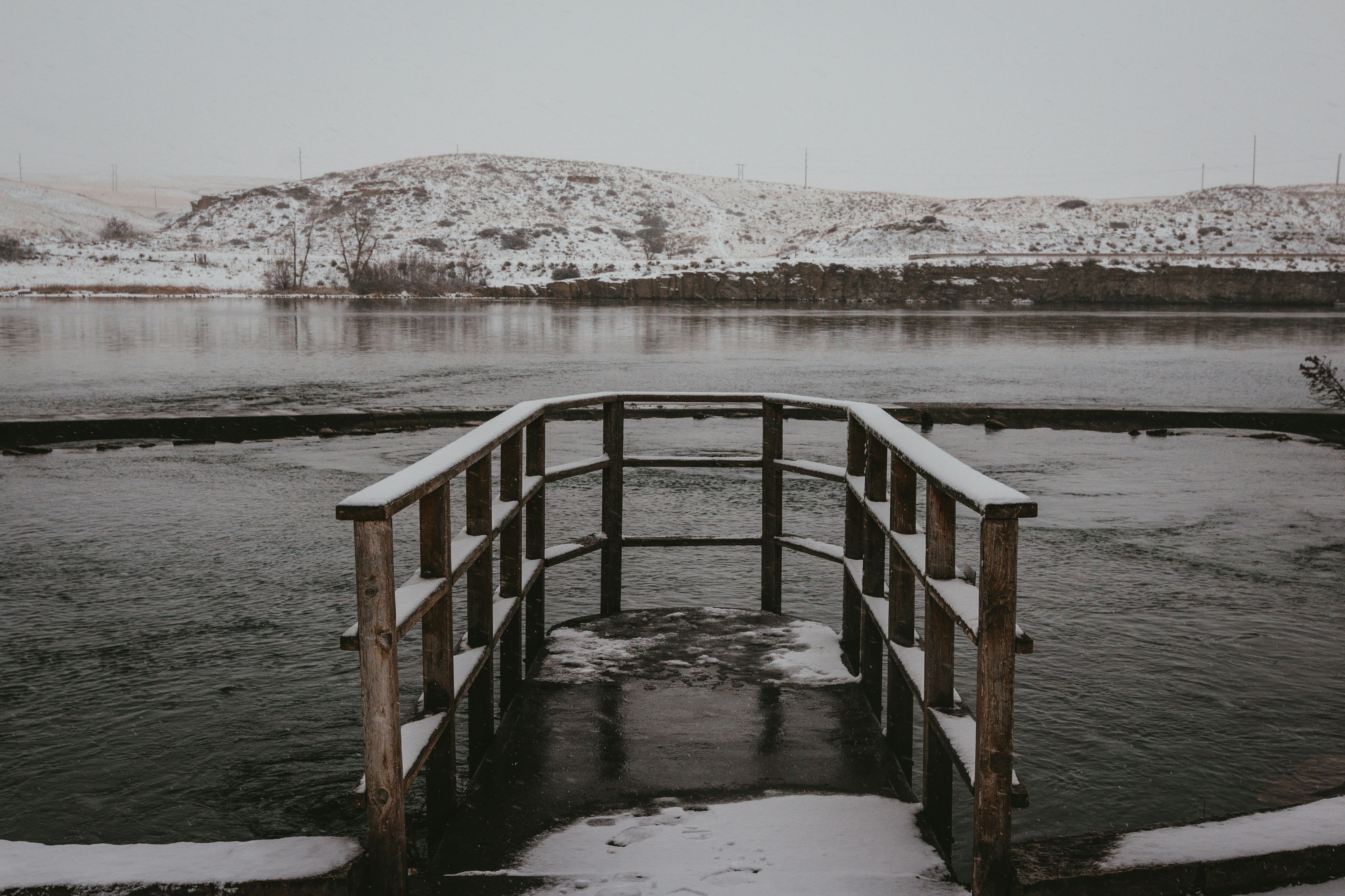 Snow-covered wooden dock over a calm, wintry lake with distant snowy hills.