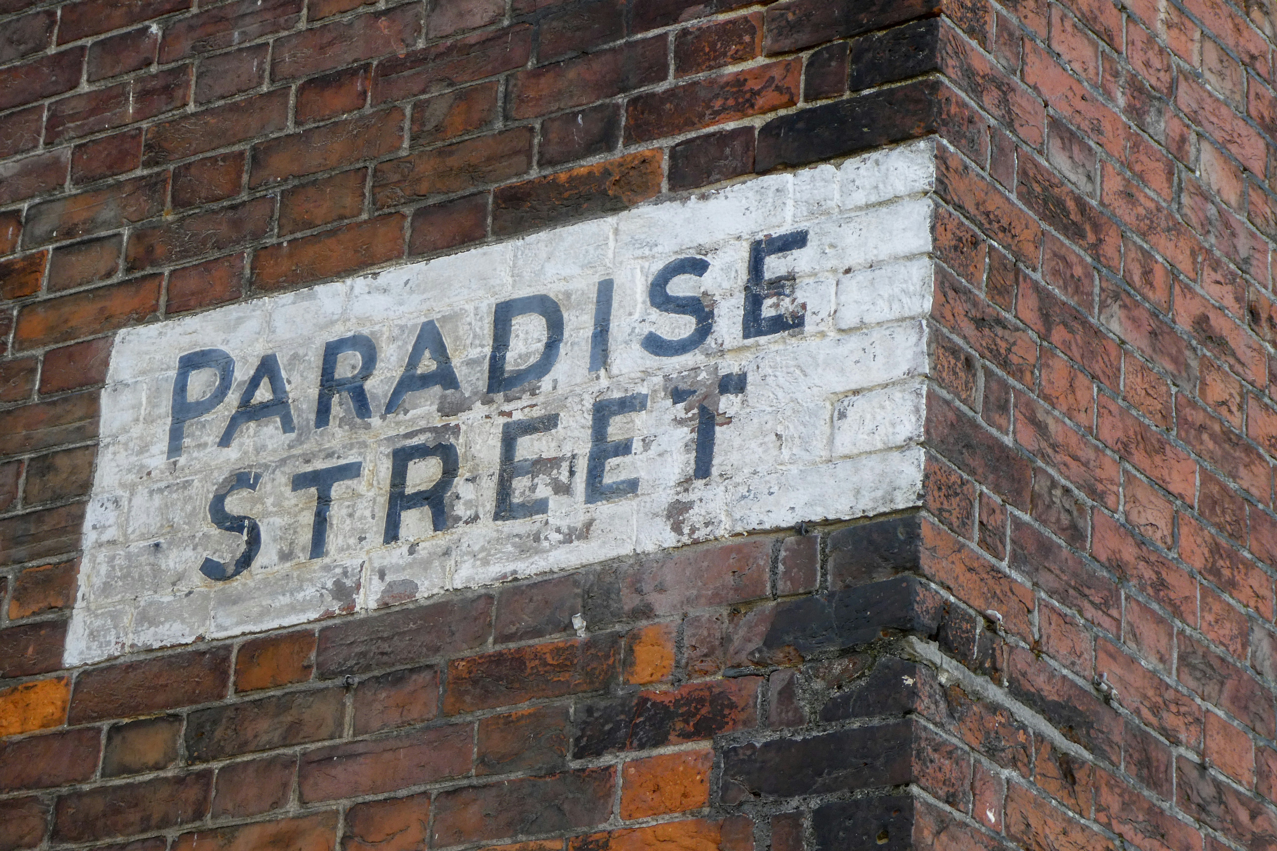 Weathered white street sign reading 'Paradise Street' mounted on a brick corner with aged mortar and varied brick tones.
