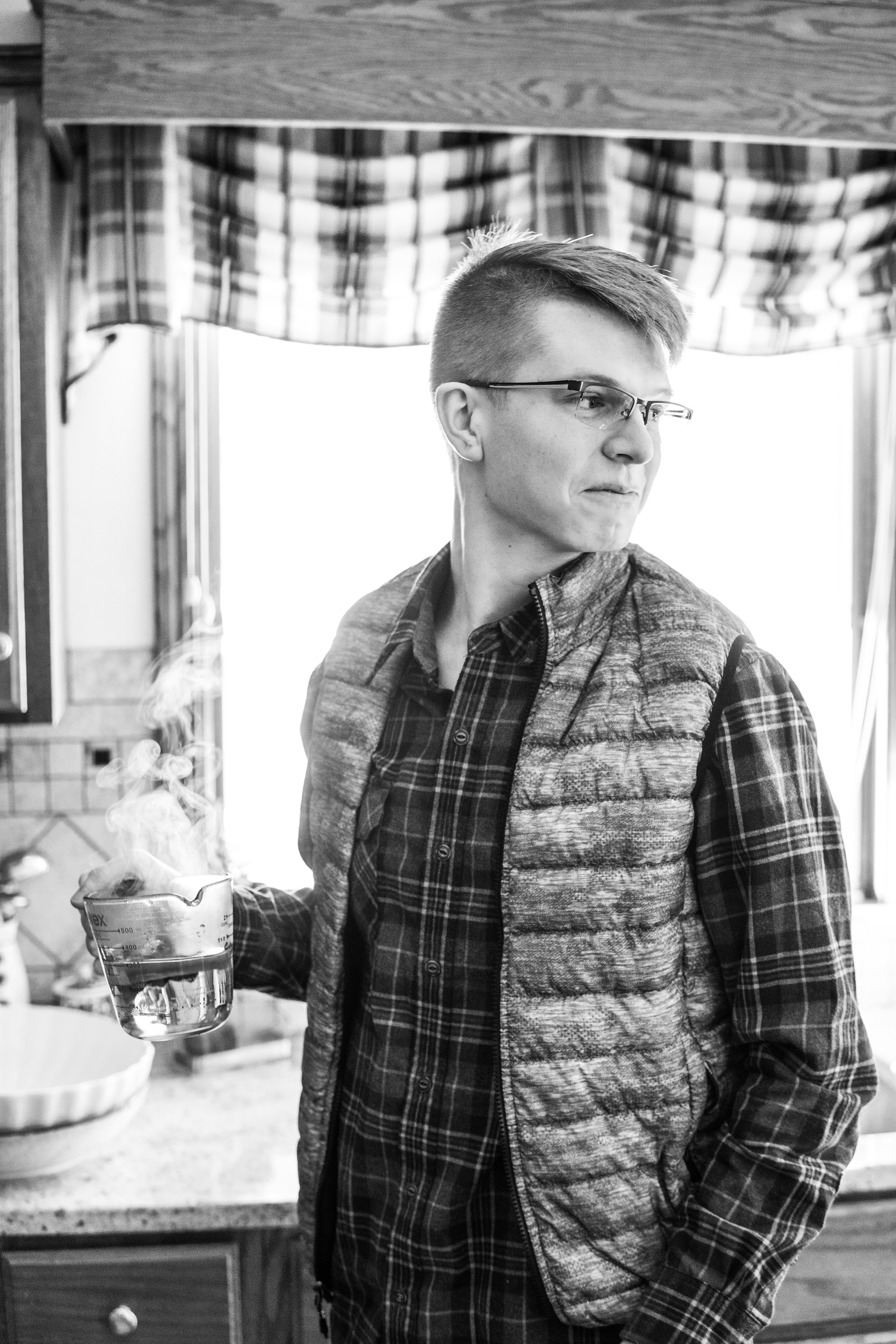 Young man in a plaid shirt and vest holding a glass with steam rising, standing in a cozy kitchen setting.