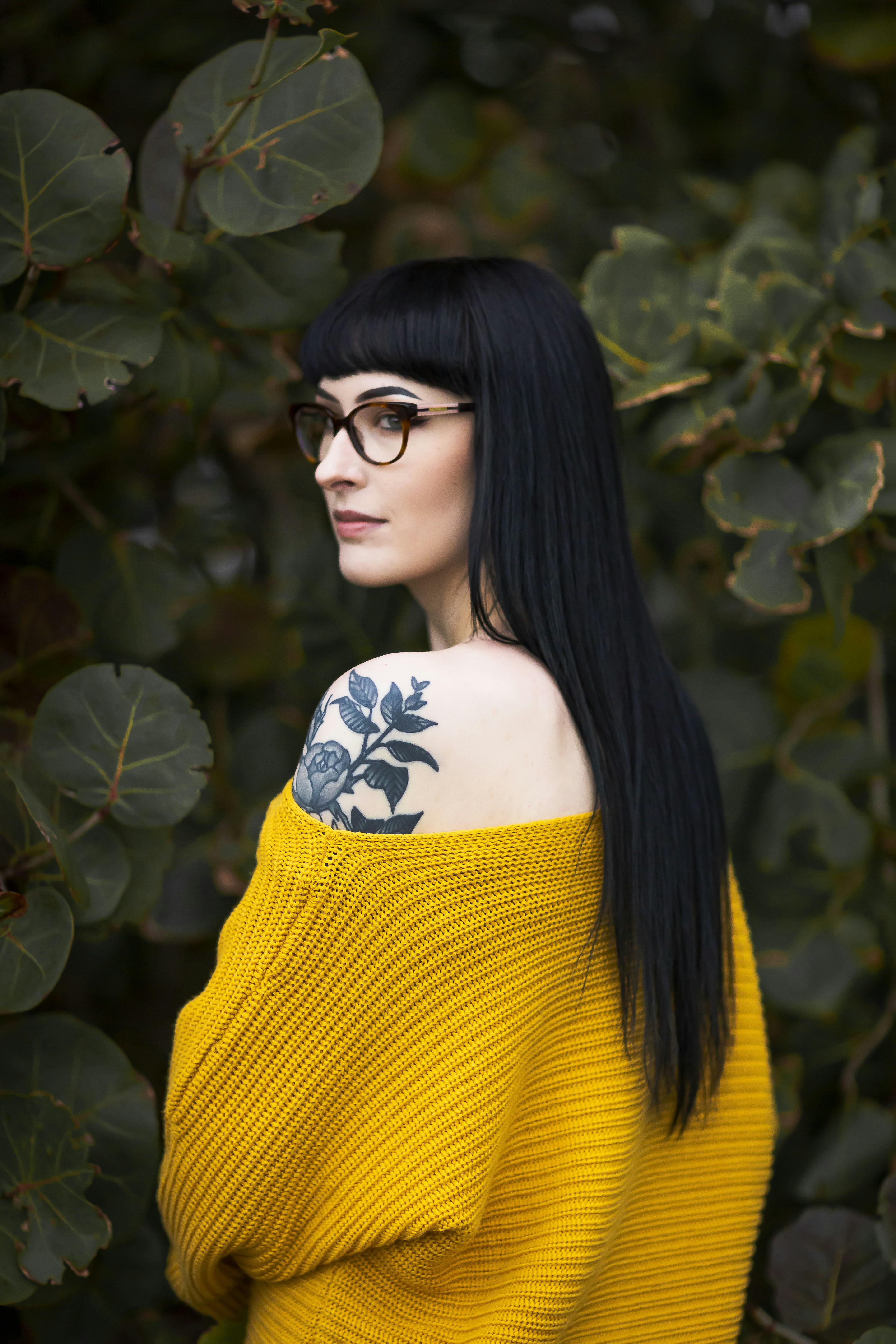 A woman with long black hair and glasses gazes over her shoulder, showcasing a floral tattoo on her bare shoulder, framed by lush green foliage. 