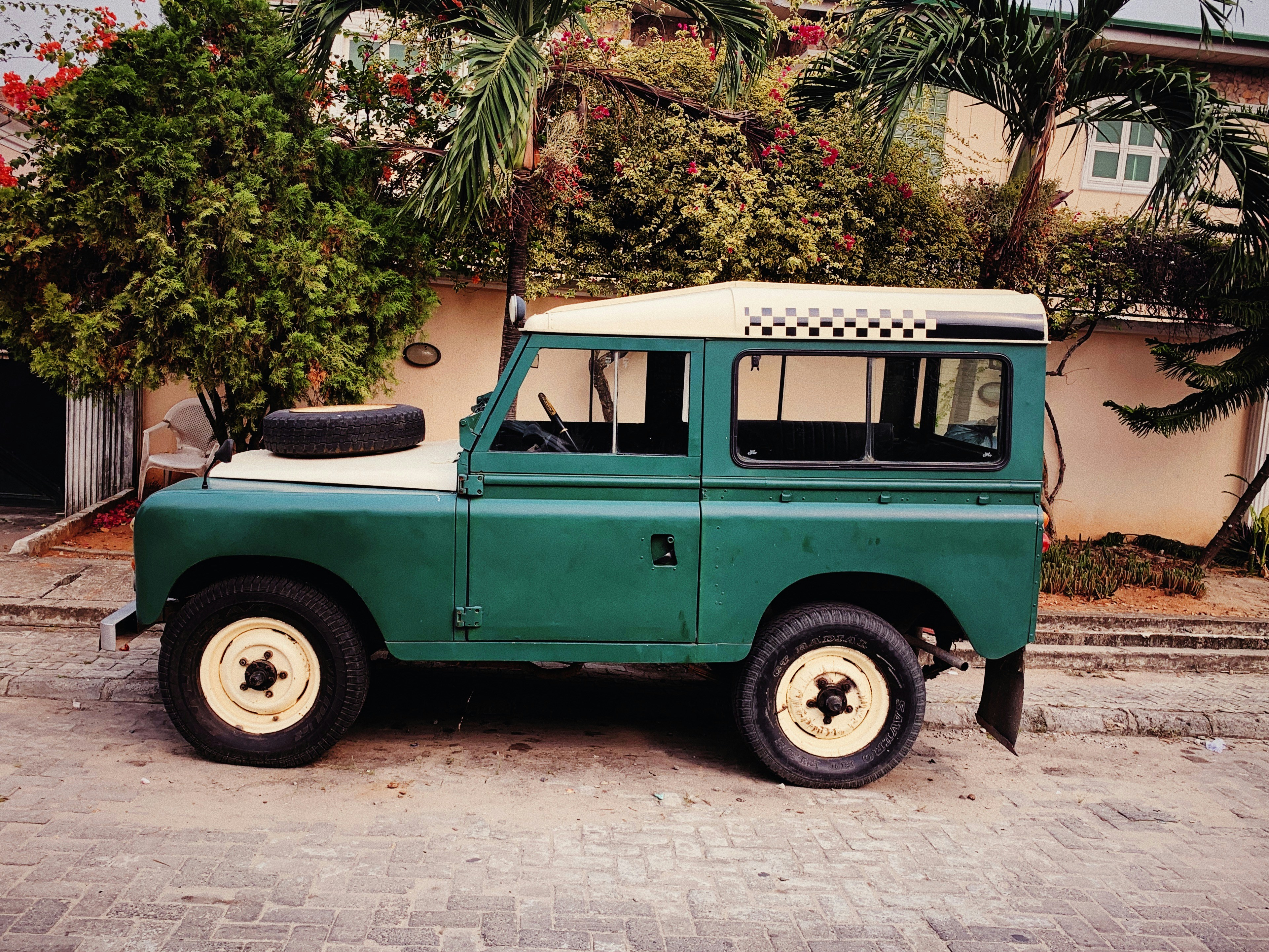 Classic green Land Rover parked on a cobblestone street, surrounded by lush foliage and vibrant flowers.