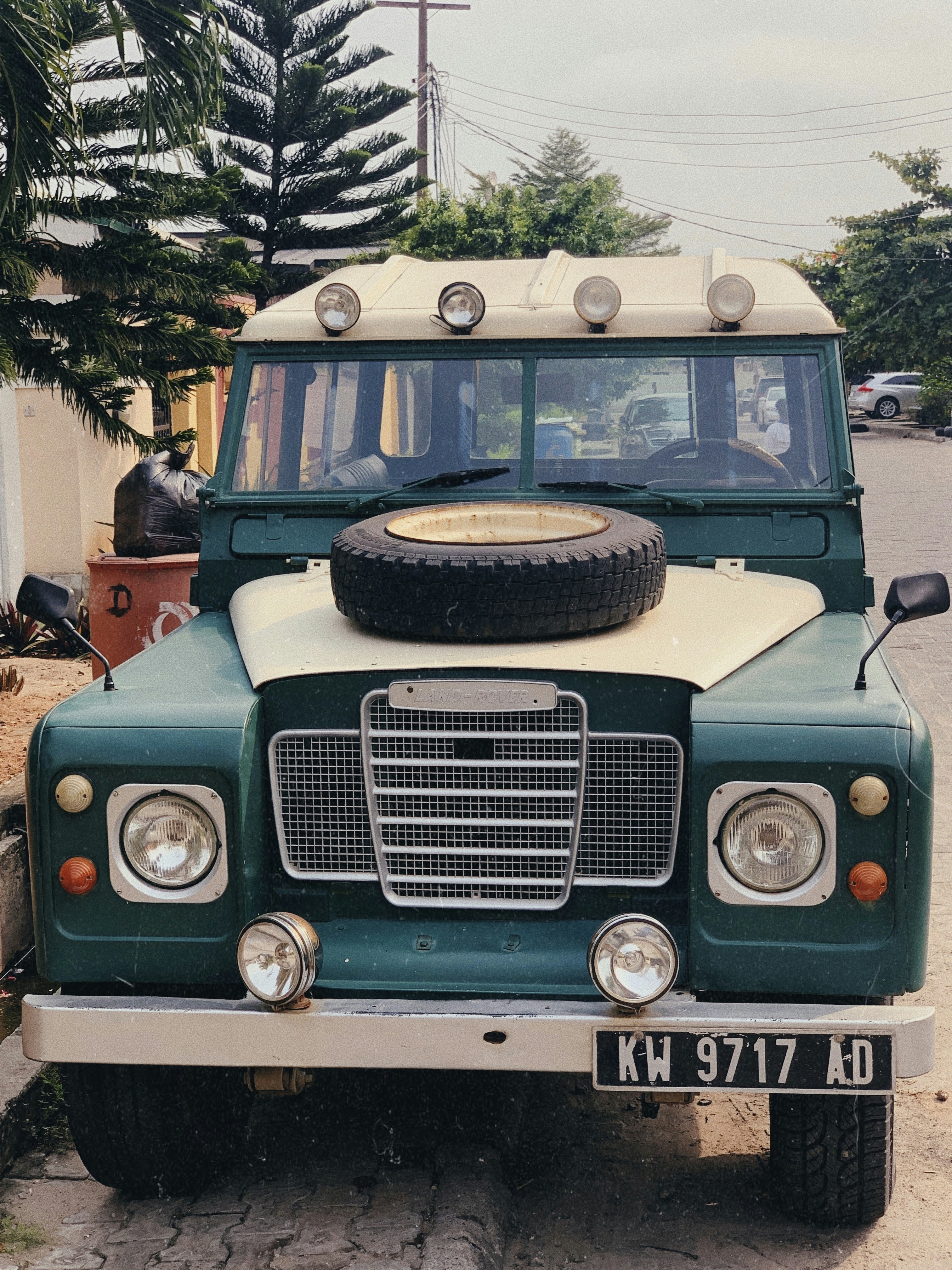 Vintage green vehicle parked on a street, featuring a spare tire on the hood and surrounded by lush foliage. The scene captures a nostalgic automotive aesthetic.