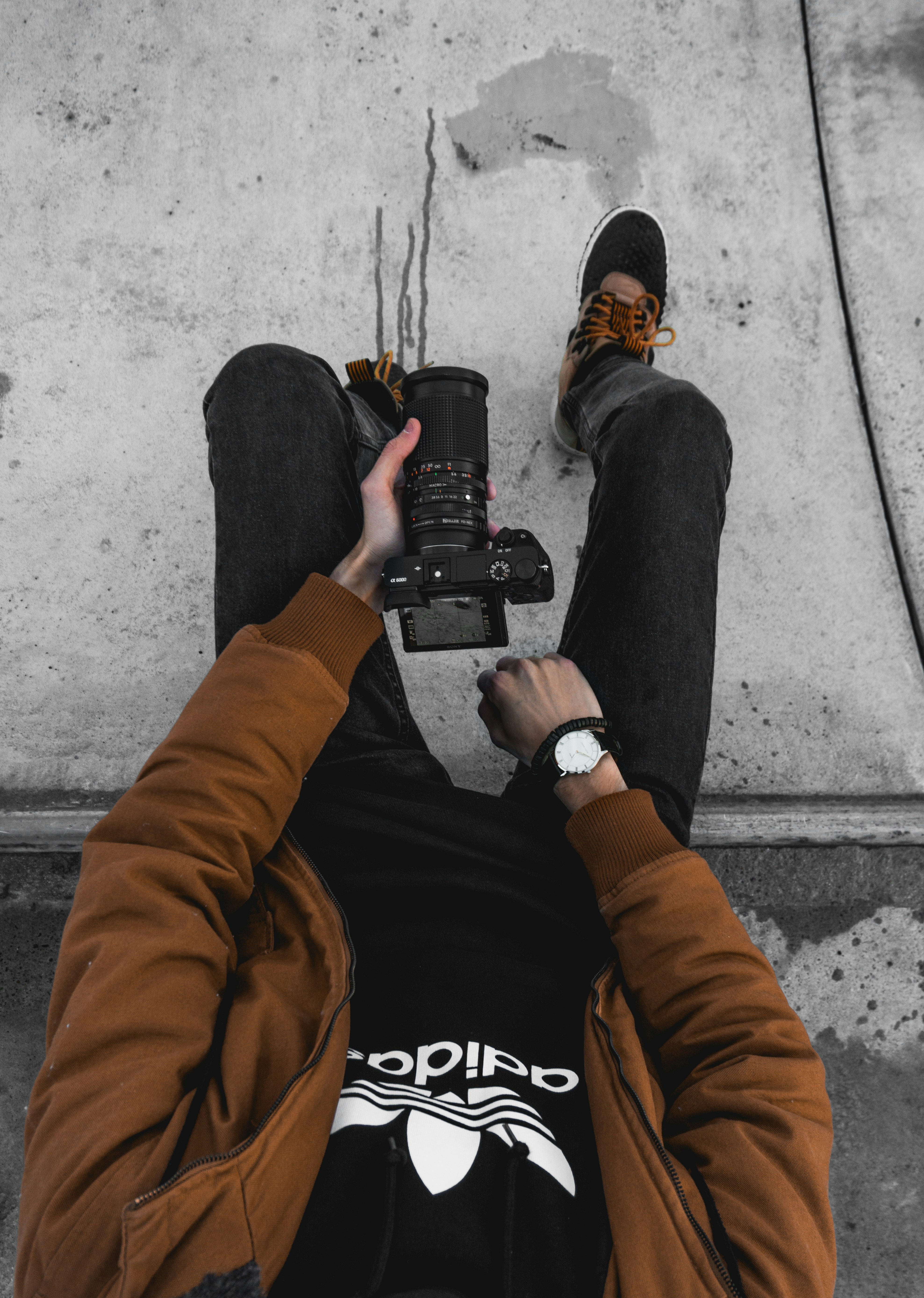 A person seated on the ground, holding a camera with a large lens, dressed in casual attire, showcasing a stylish watch. The scene captures the essence of photography culture.