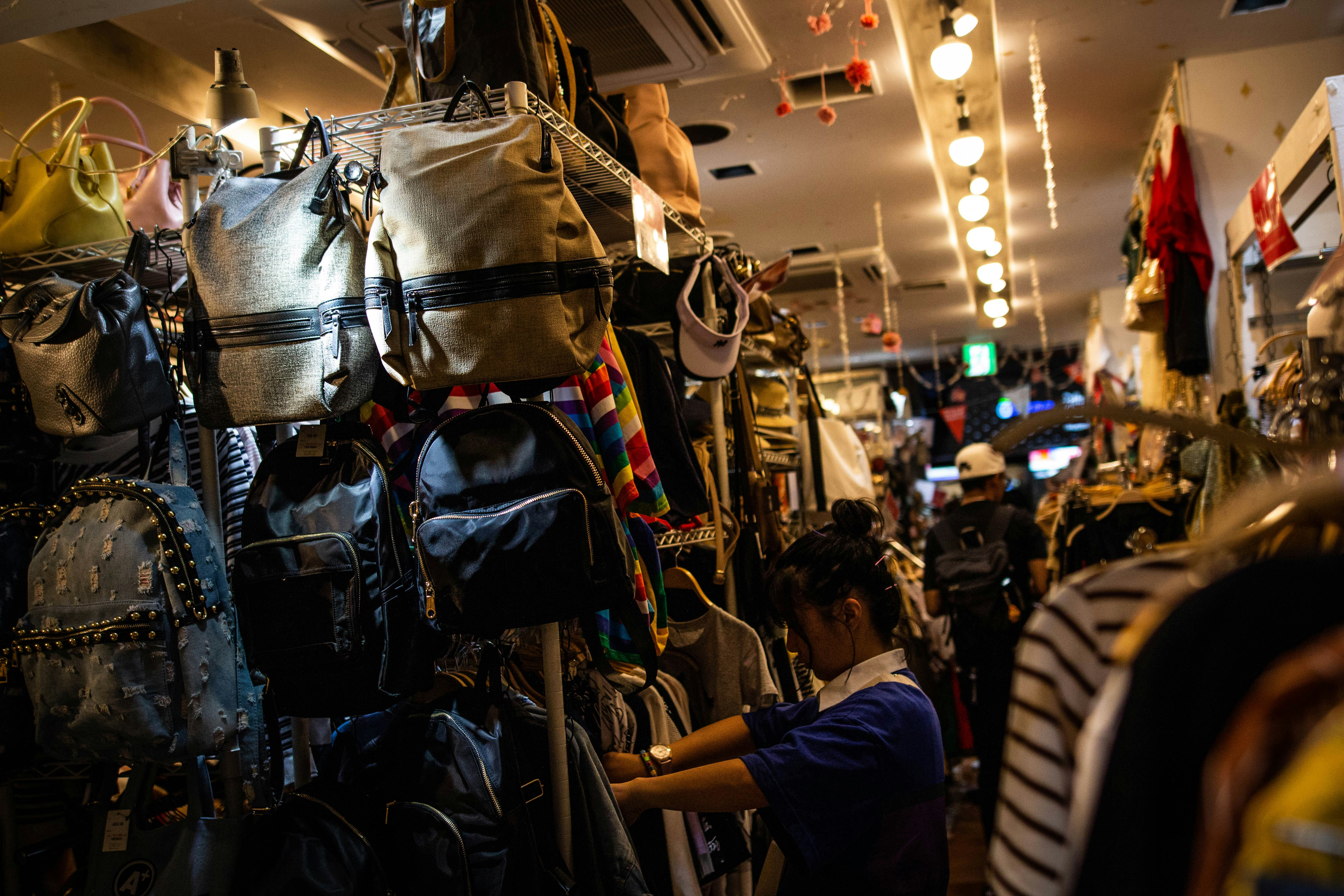 A bustling retail space filled with an array of bags and accessories, showcasing vibrant colors and diverse styles. A shopper browses among the crowded displays.