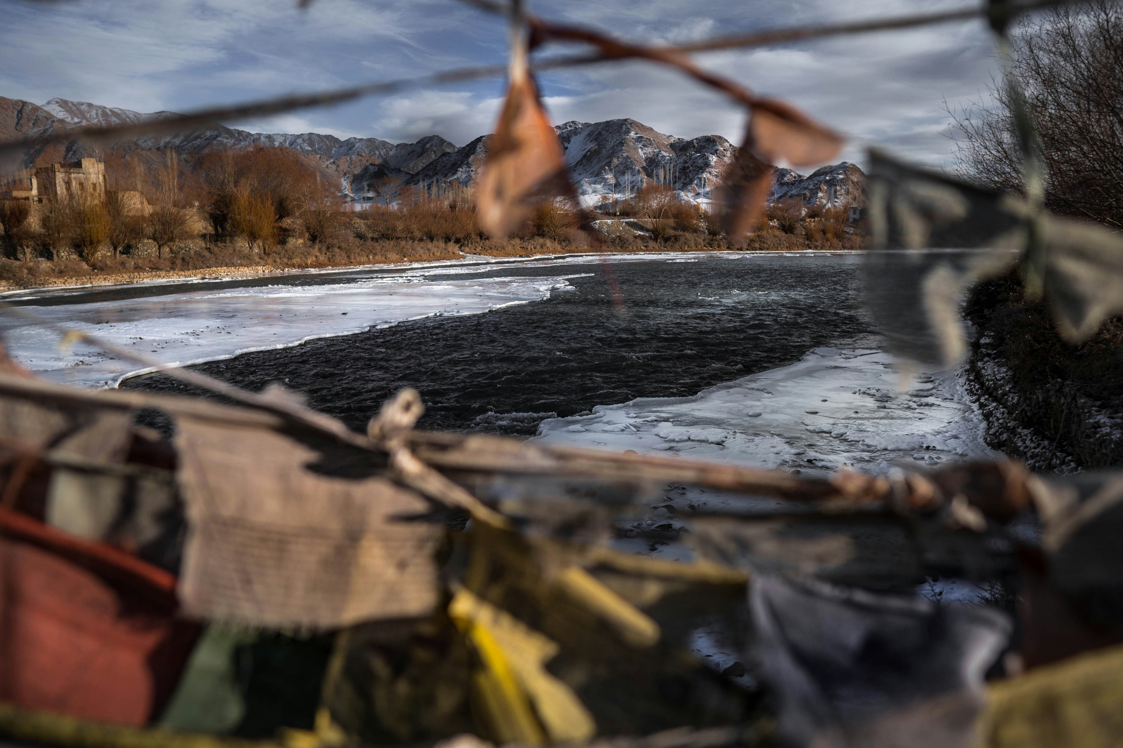 Colorful prayer flags flutter above a frozen river with snow-capped mountains in the background.