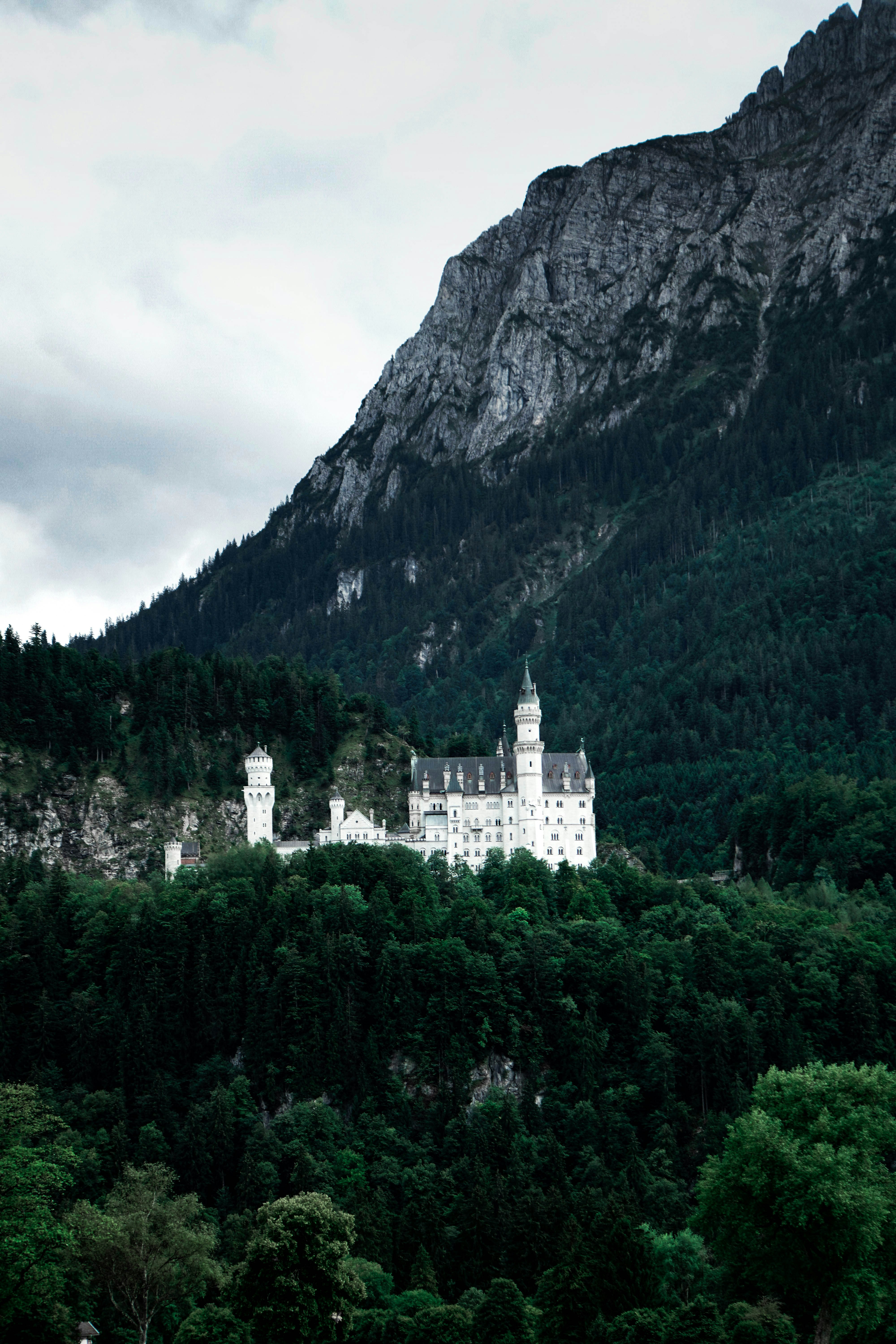 A majestic castle nestled among lush greenery, framed by towering mountains under a moody sky.