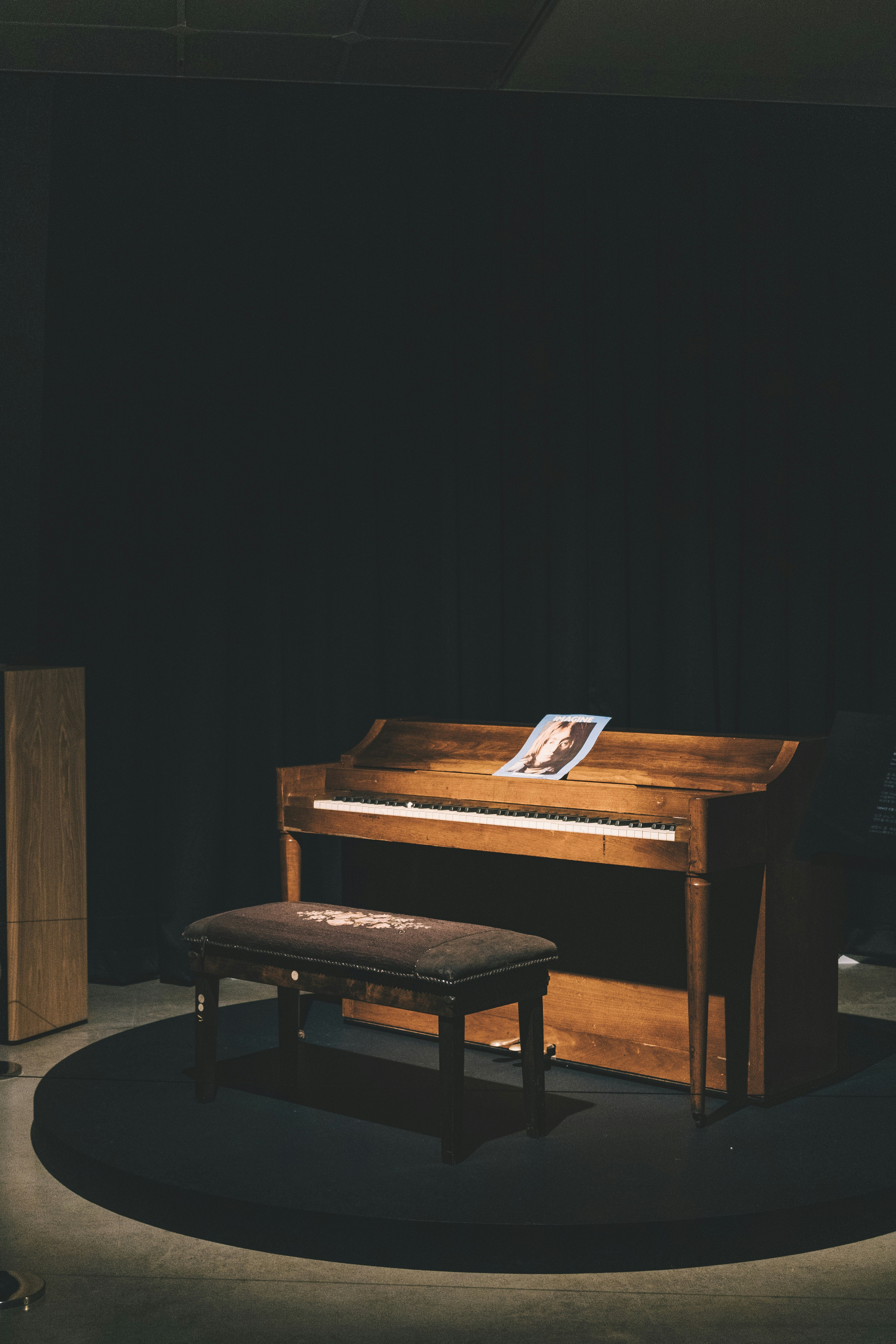 A wooden piano with a sheet of music rests on a circular platform, illuminated against a dark backdrop. A plush bench complements the scene.