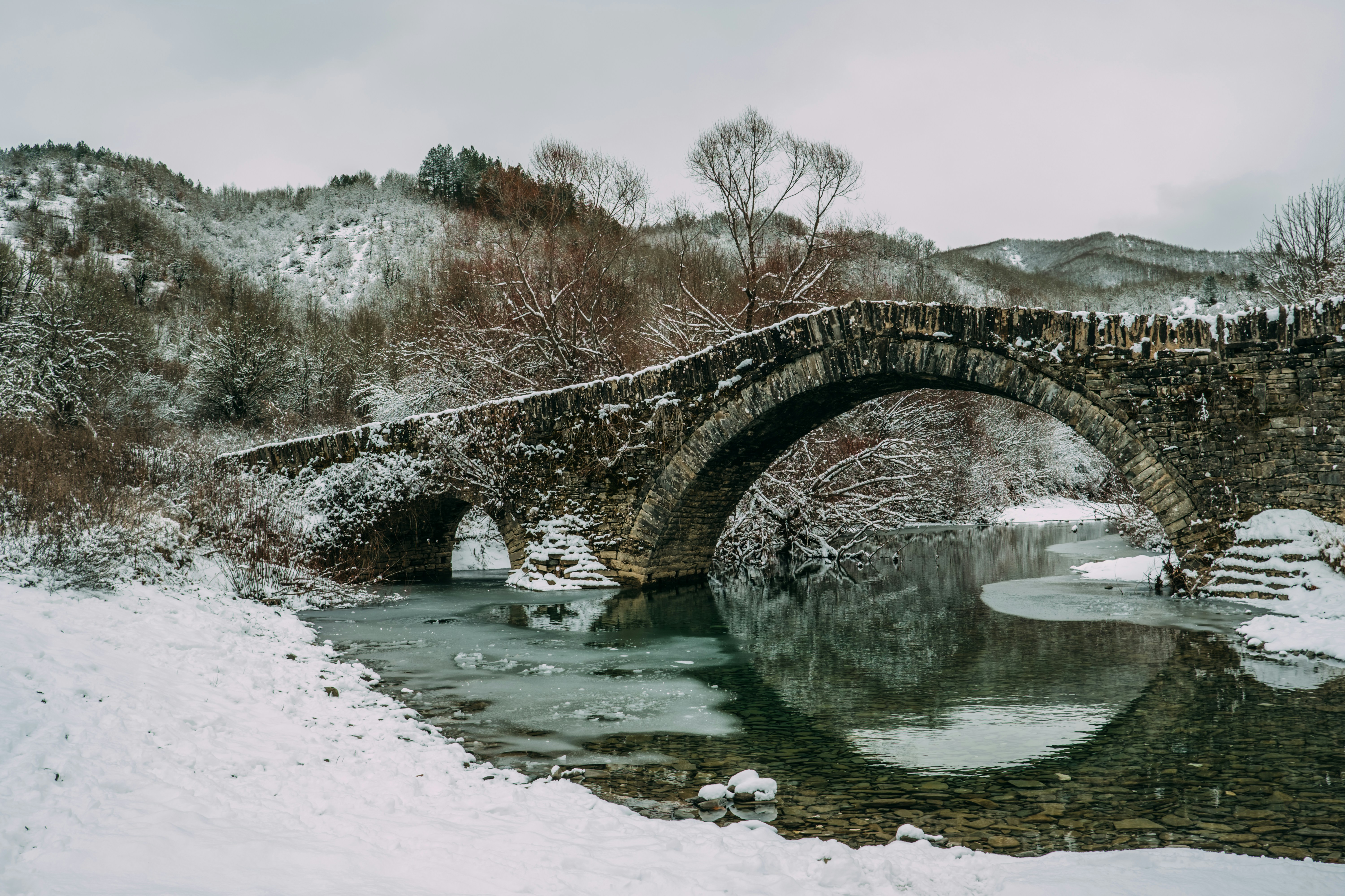 Ancient stone bridge arching over a serene, snow-covered river, surrounded by winter trees. Tranquil scene capturing the essence of a quiet, frosty landscape.