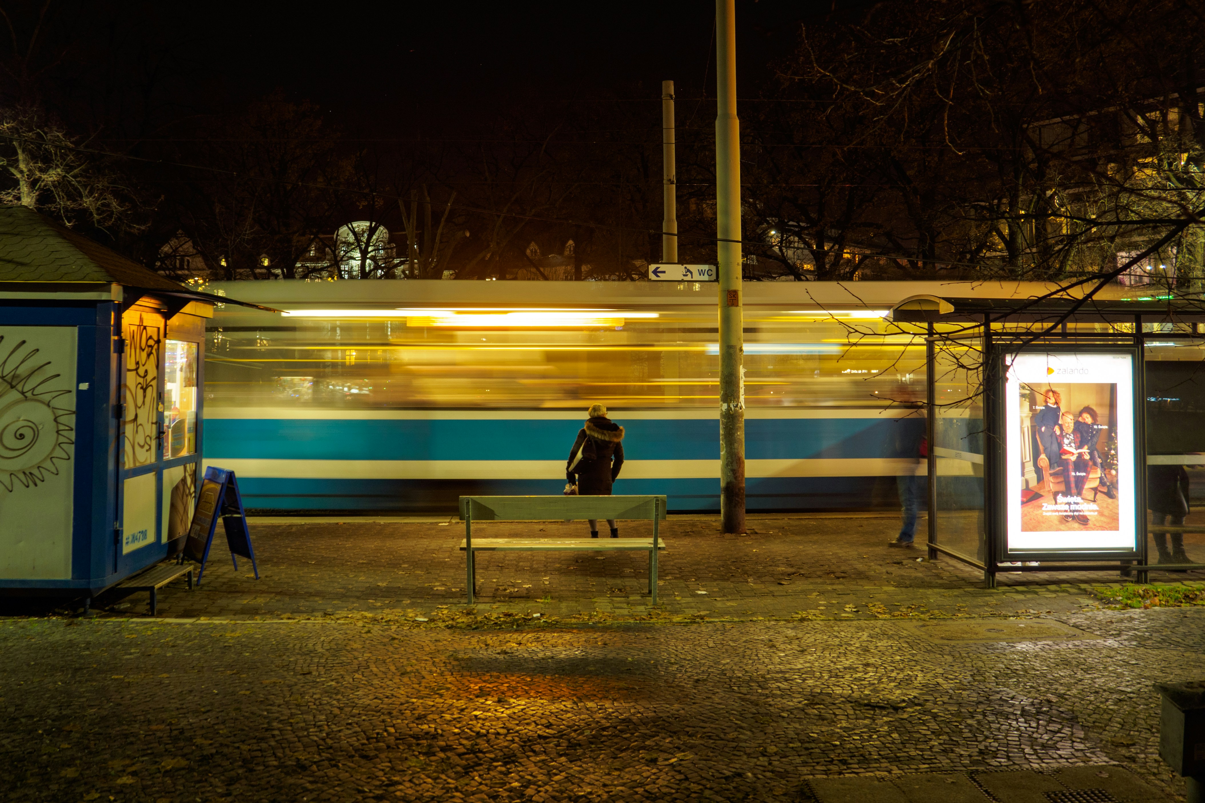 Person standing near a brightly lit tram station with a passing train in motion at night.