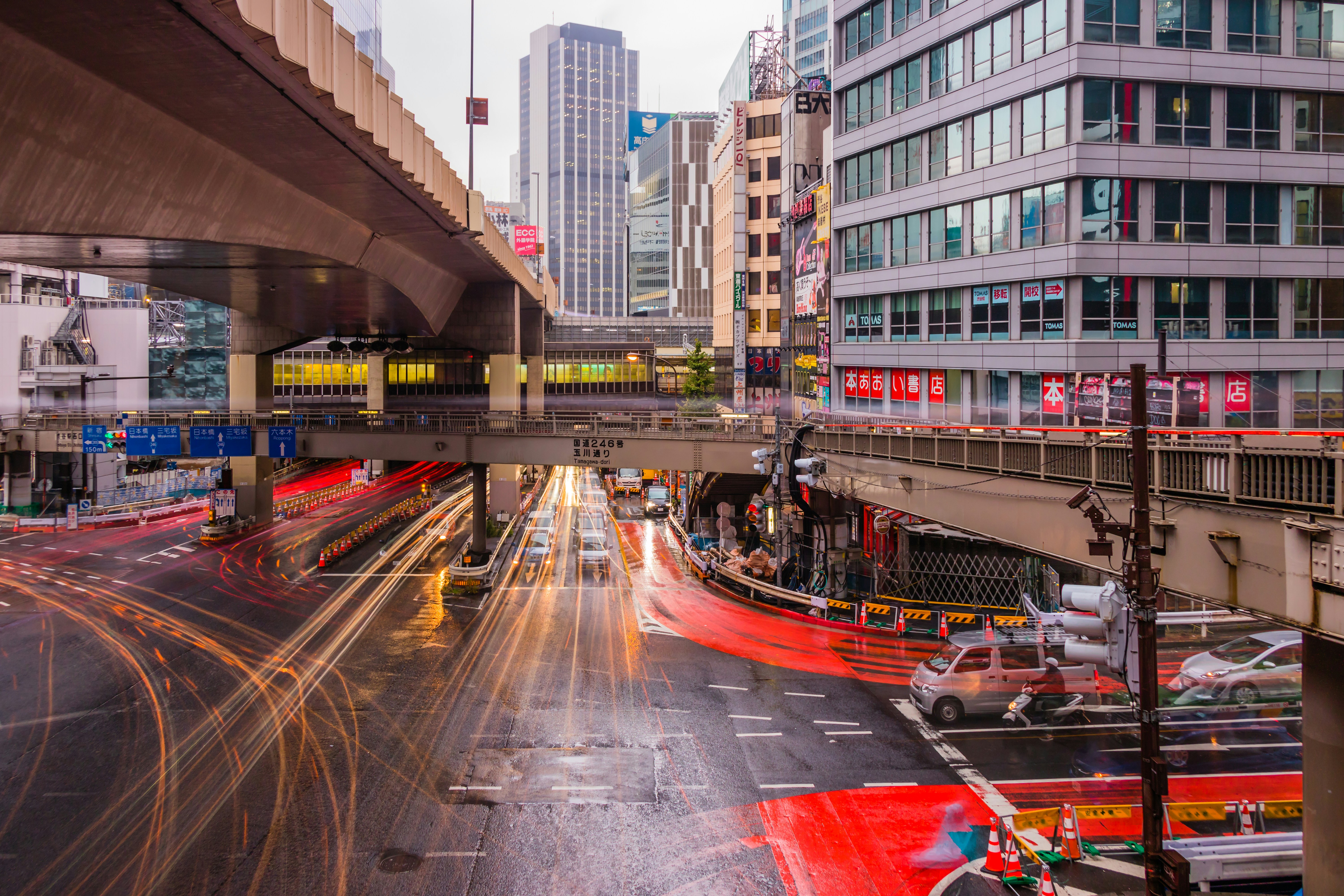 City intersection with streaking car lights beneath towering skyscrapers and elevated highways at dusk.