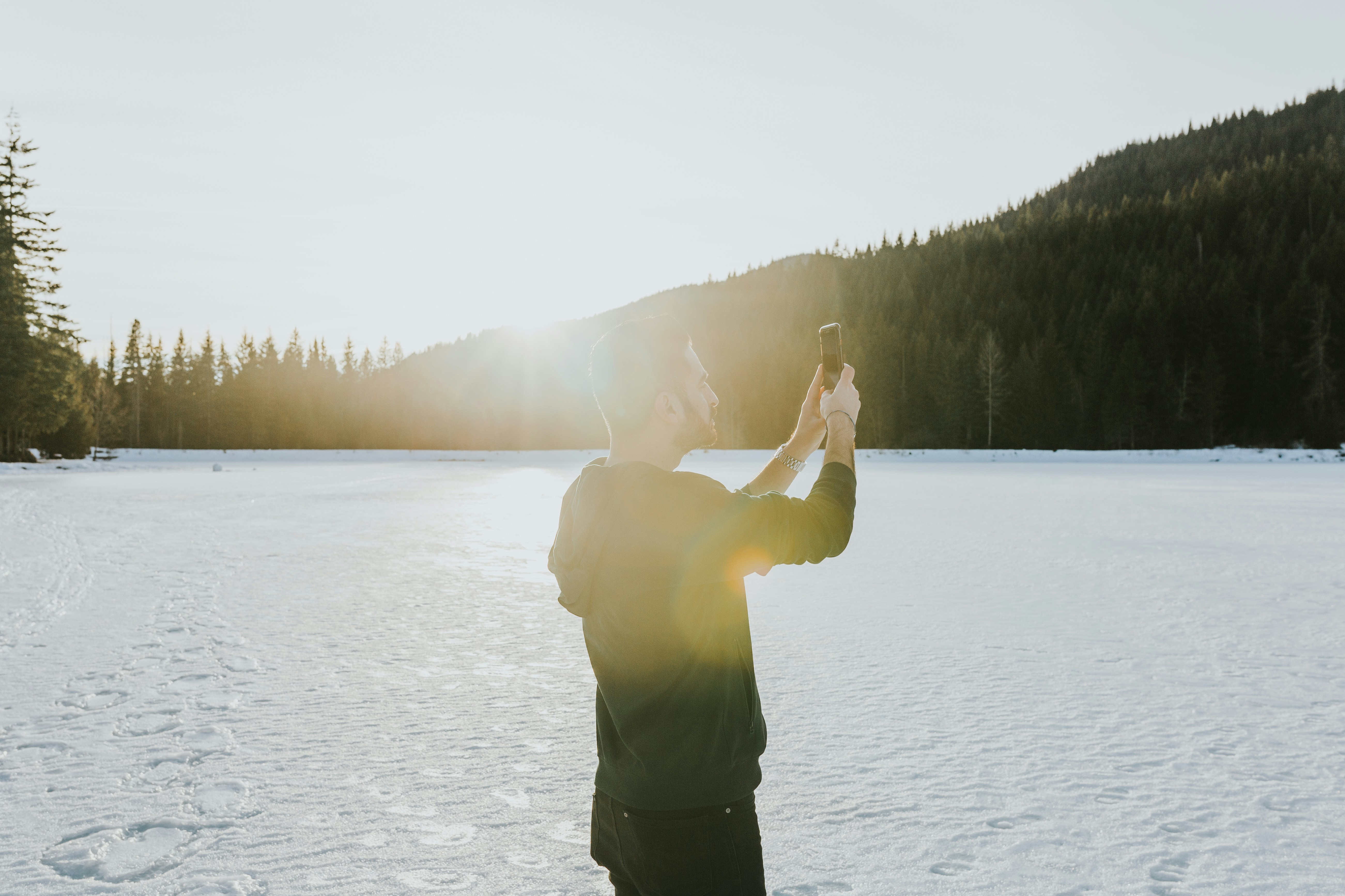 Person capturing a moment on their phone against a backdrop of a snowy lake and mountains, illuminated by the sun's rays.