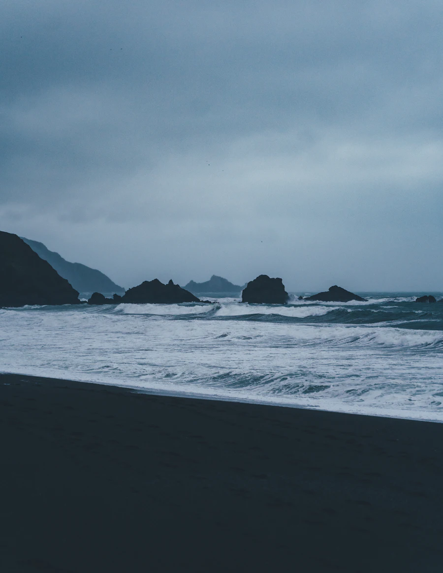 Moody seascape with dark sand beach, white surf, and jagged sea stacks under a heavy overcast sky. Distant cliffs fade into mist on the left.