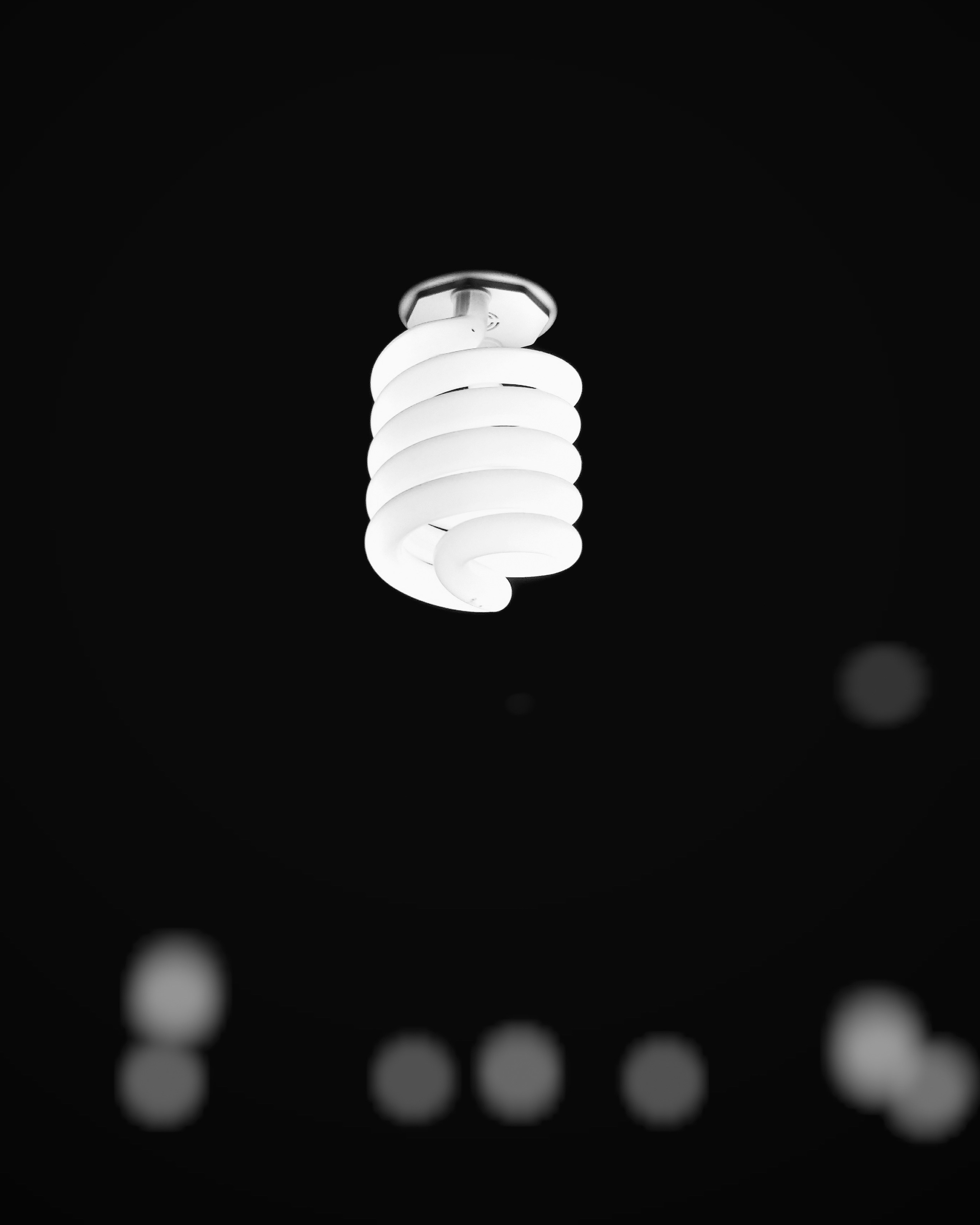 Spiral-shaped energy-saving bulb suspended against a dark background, creating a striking contrast. Soft bokeh effects from distant light sources enhance the ambiance.