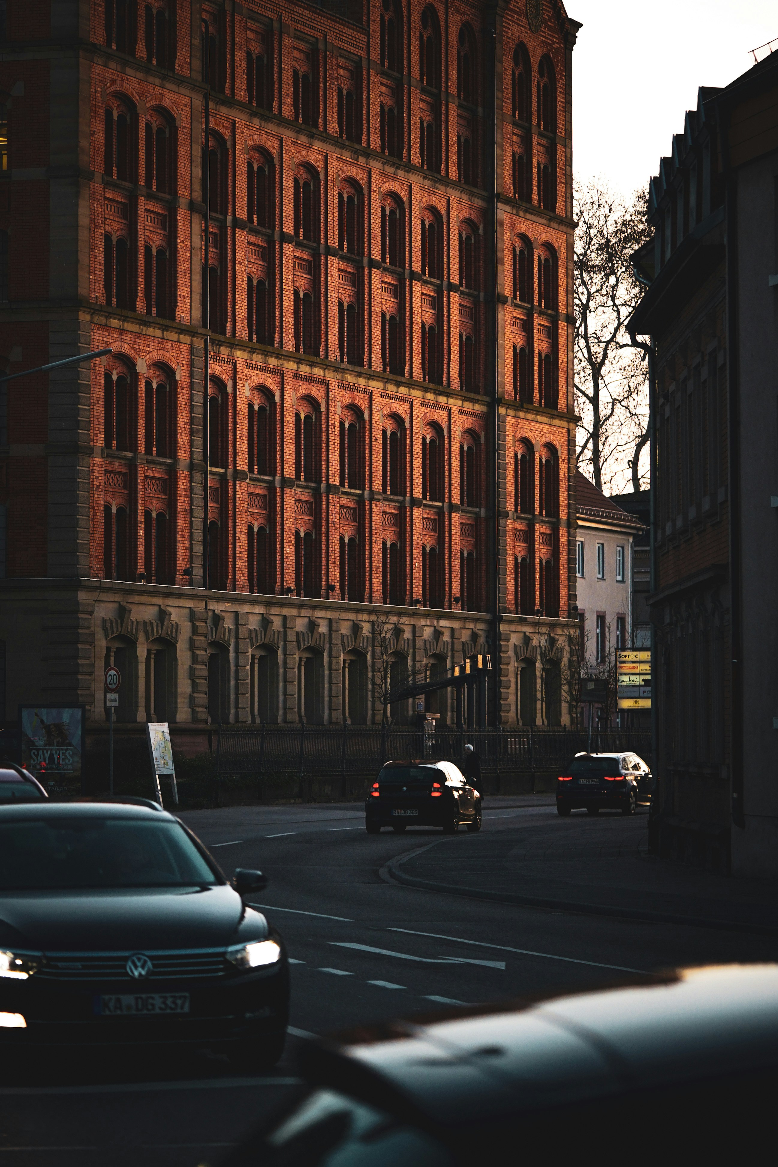 Historic brick building illuminated by warm sunset light, casting long shadows on the street. Vehicles navigate the curved road in the foreground.