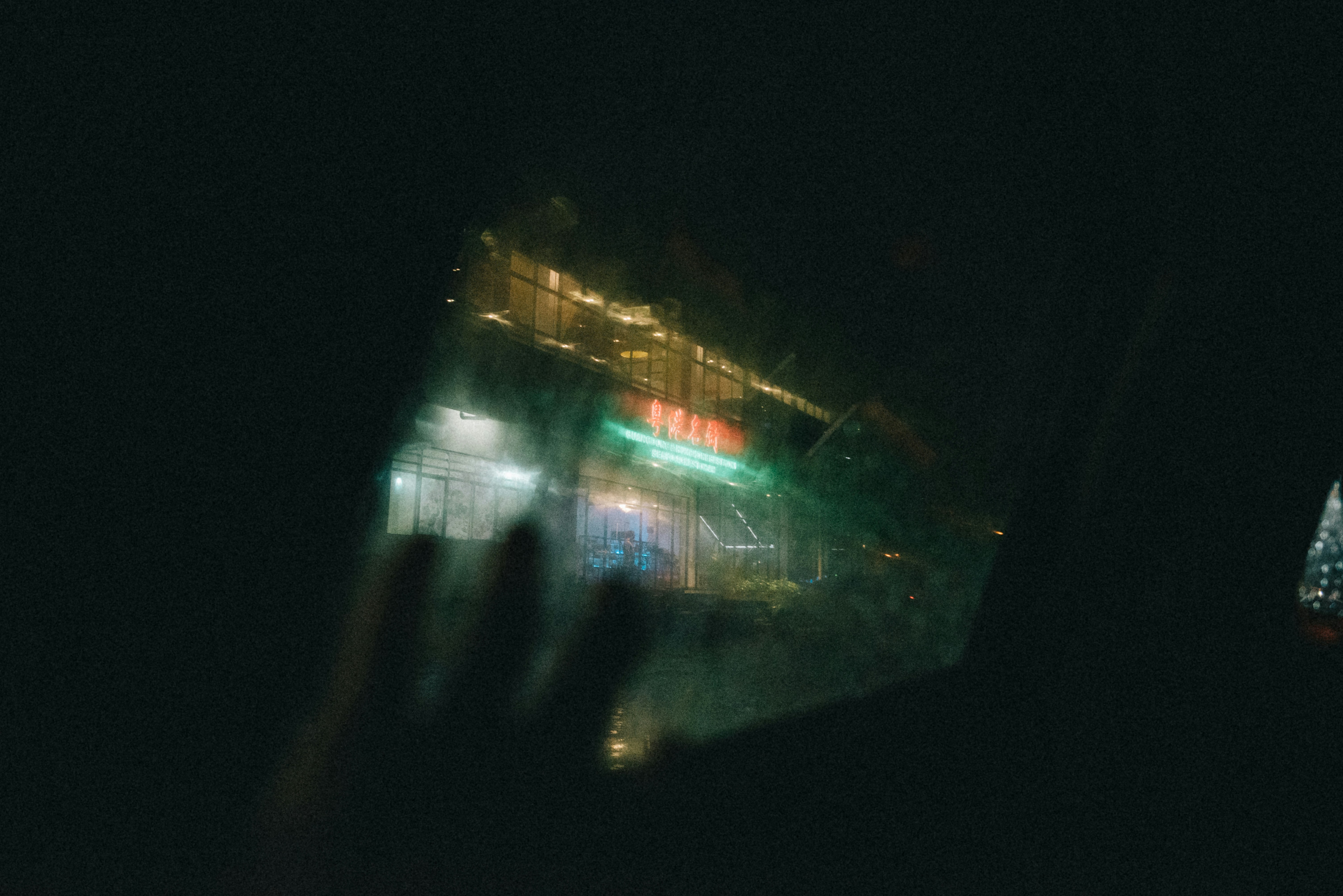 A hand reaches toward a brightly lit building, viewed through a rain-speckled window at night. The neon glow contrasts with the dark ambiance.