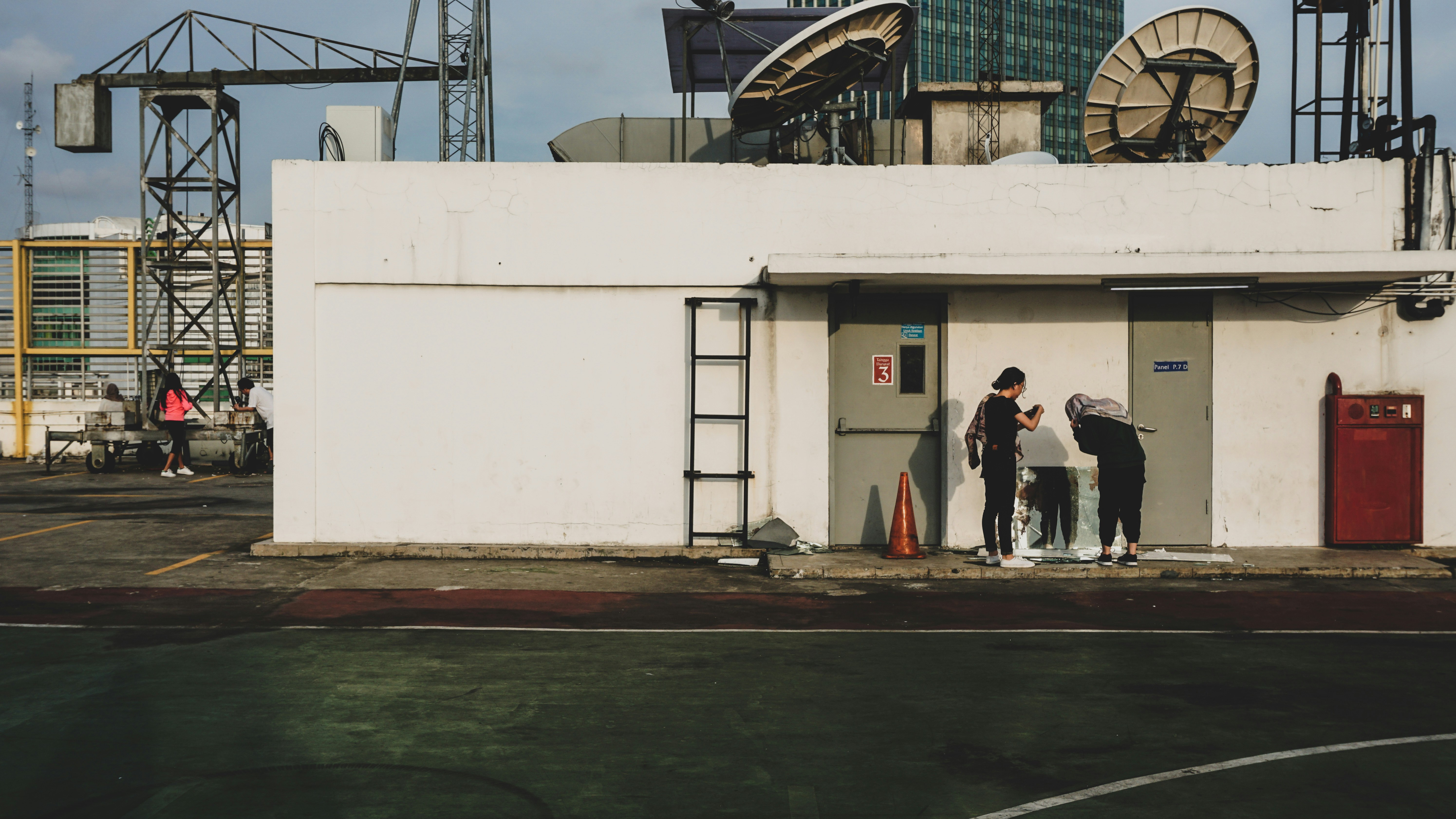 Two individuals engaged in conversation near a white structure on a rooftop surrounded by industrial elements. The scene captures a blend of urban life and architecture.