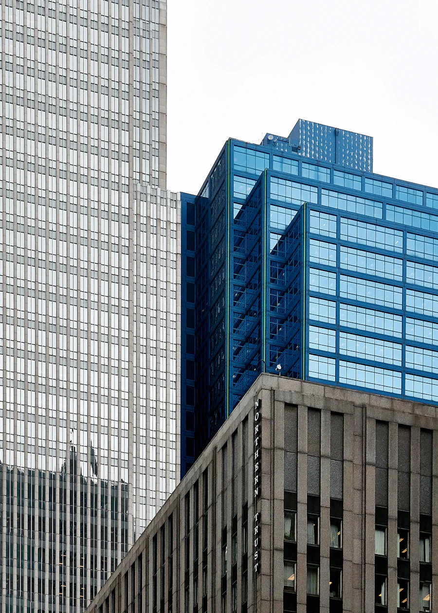 Tall modern office buildings with a blue glass tower framed by a pale sky and a gray high-rise in the foreground. Vertical lines and reflective windows create a dense urban skyline.