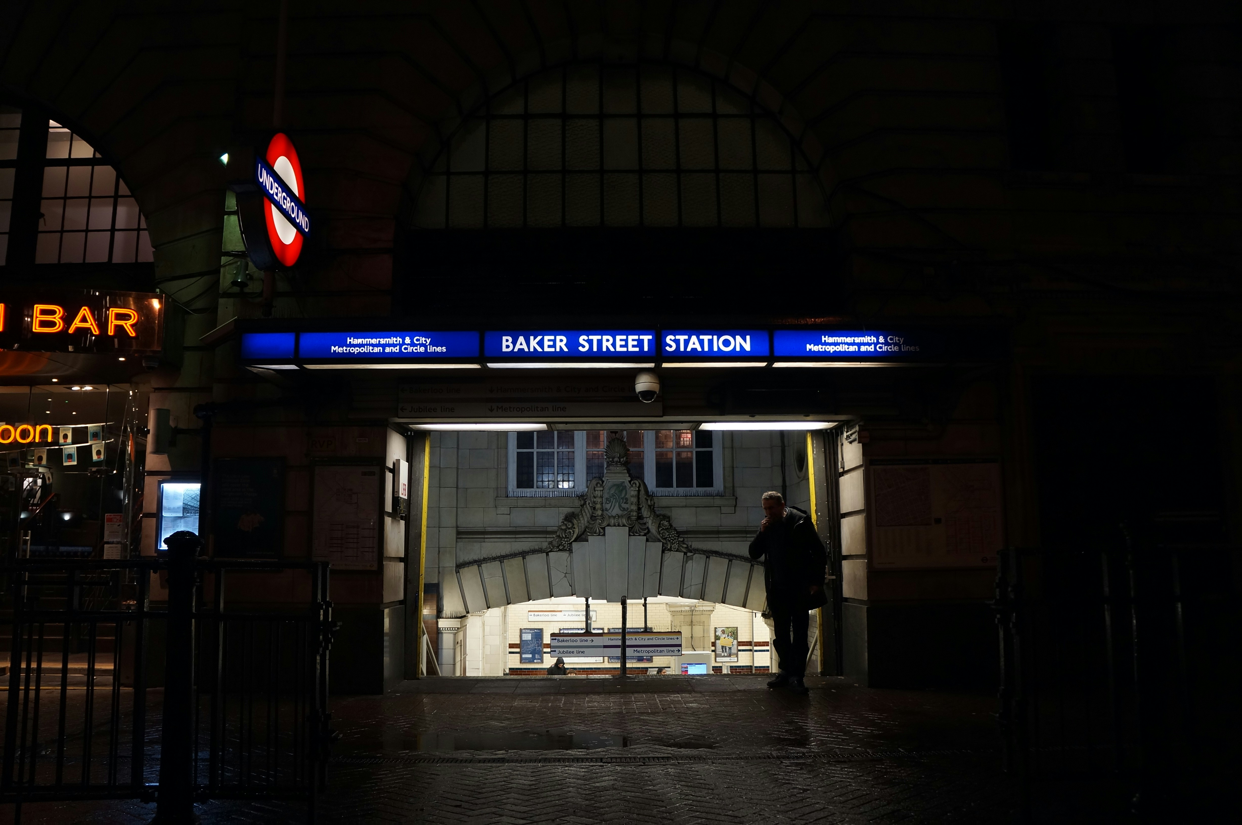 Illuminated entrance of Baker Street Station at night, showcasing its iconic signage and architectural details. A lone figure stands at the threshold.